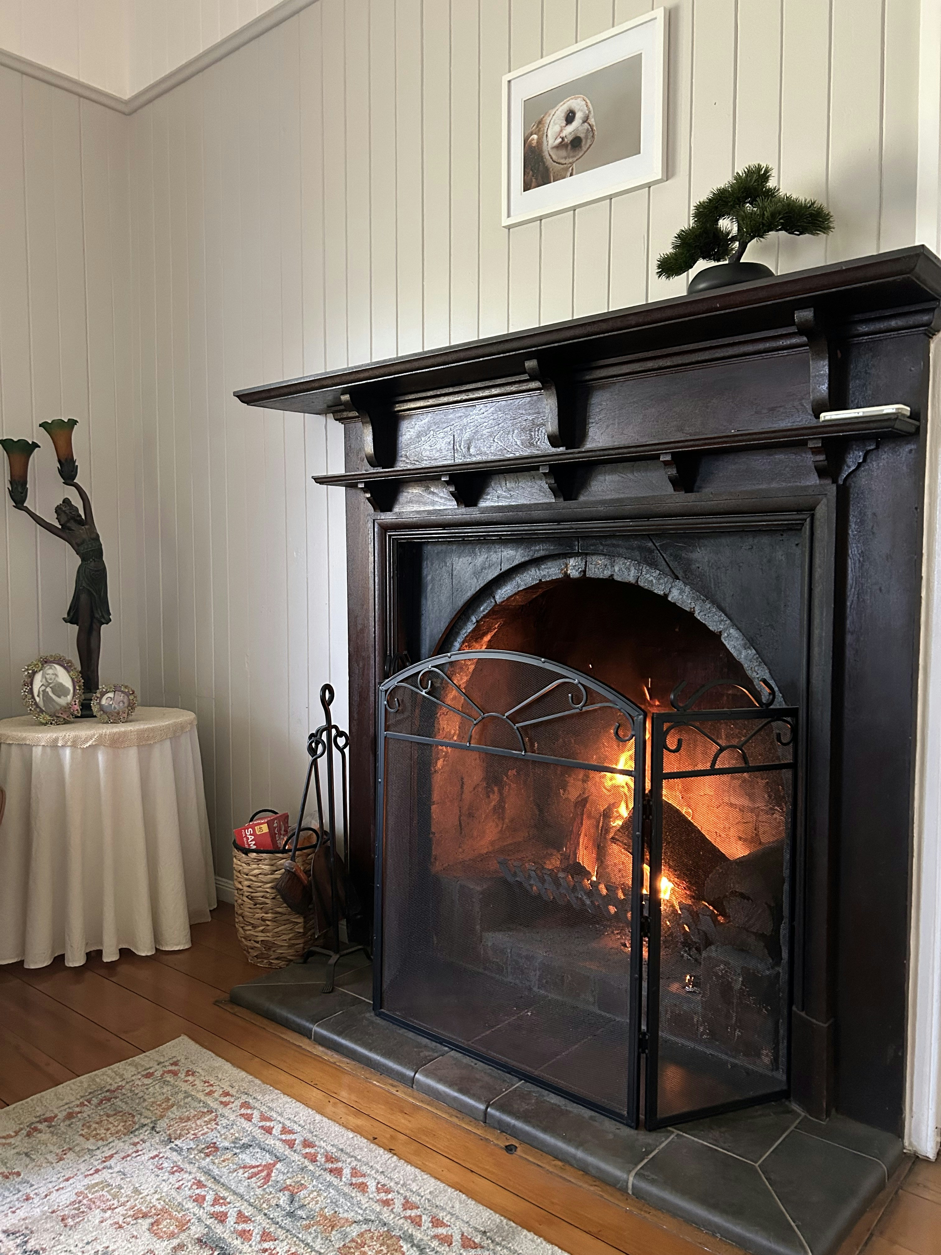 Cozy living room fireplace with a wrought-iron screen and glowing embers, dark wooden mantel, and a framed dog portrait above it.