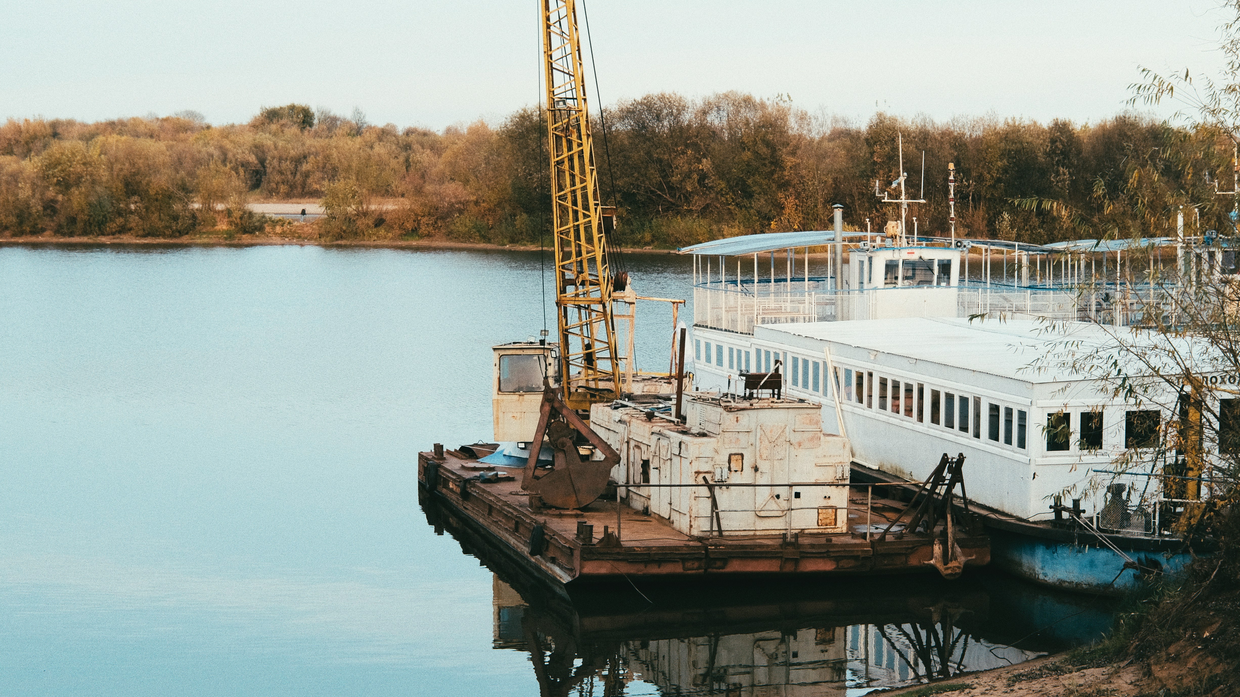 Rustic barge with crane moored on a tranquil river, surrounded by autumn trees.