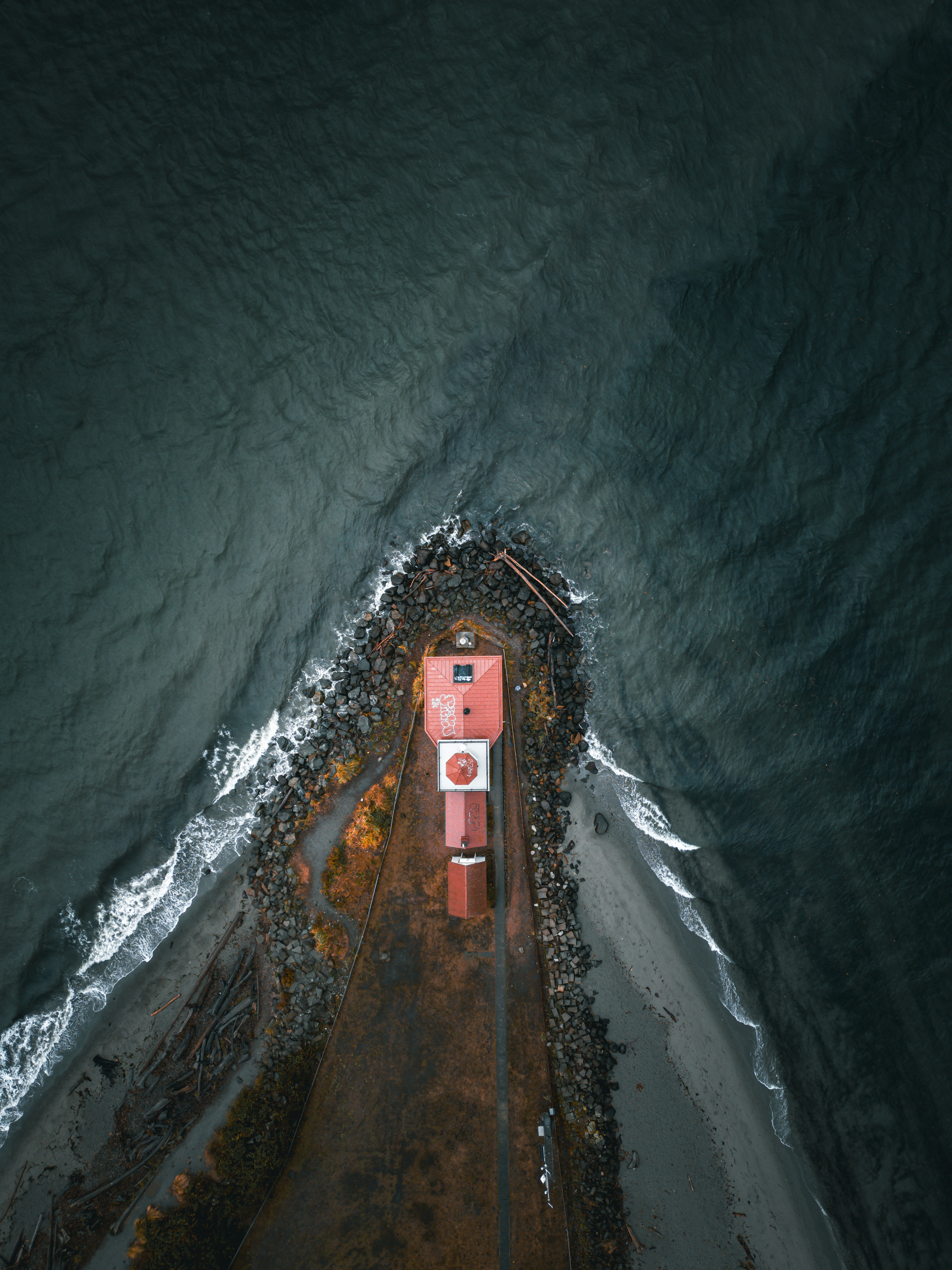 Aerial photograph of a narrow breakwater ending in a red-roofed building. The structure is bordered by dark blue sea and breaking waves along the sides.