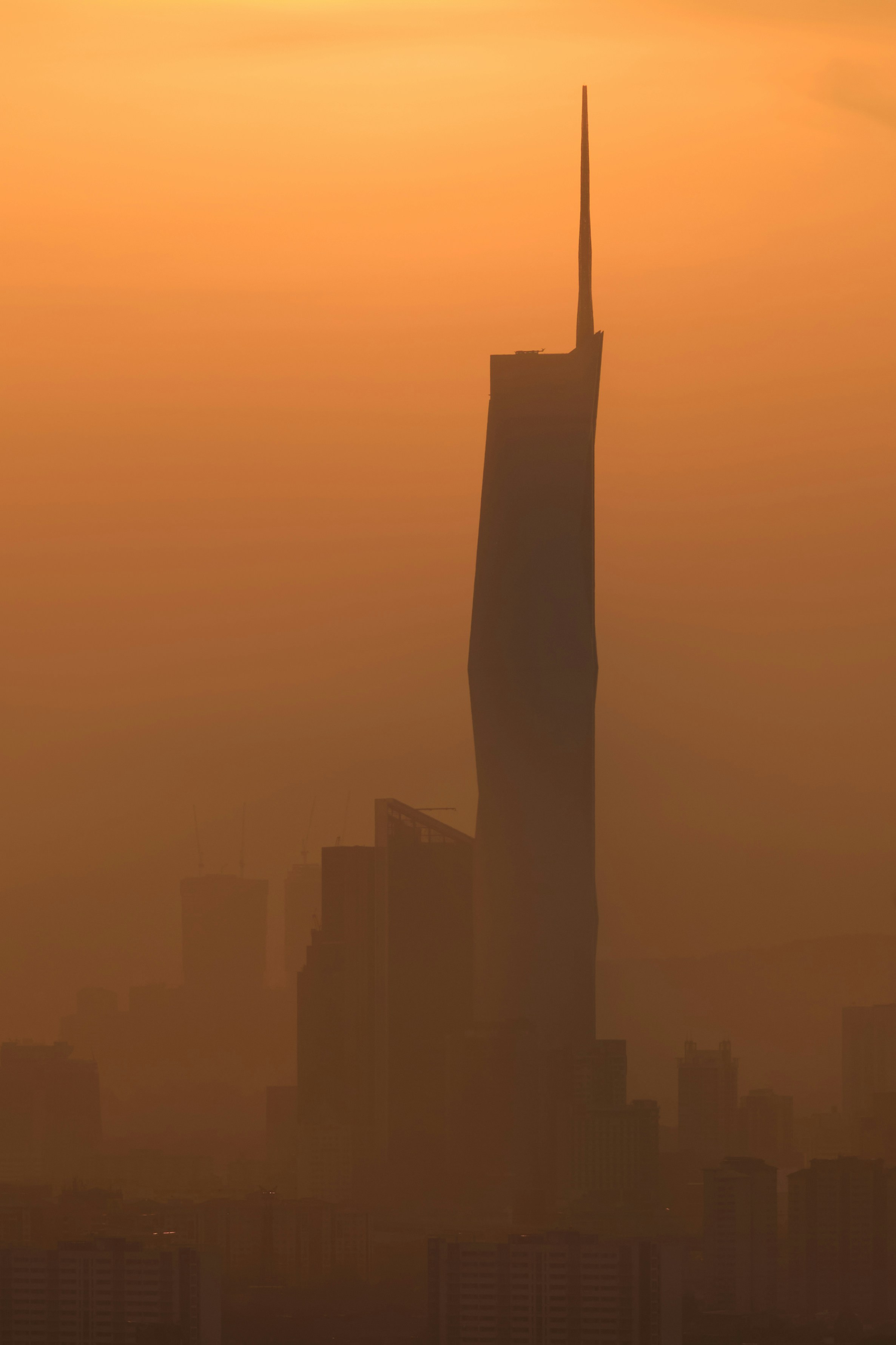 Tall, tapered skyscraper pierces the orange sunset haze, with a muted city silhouette in the foreground.