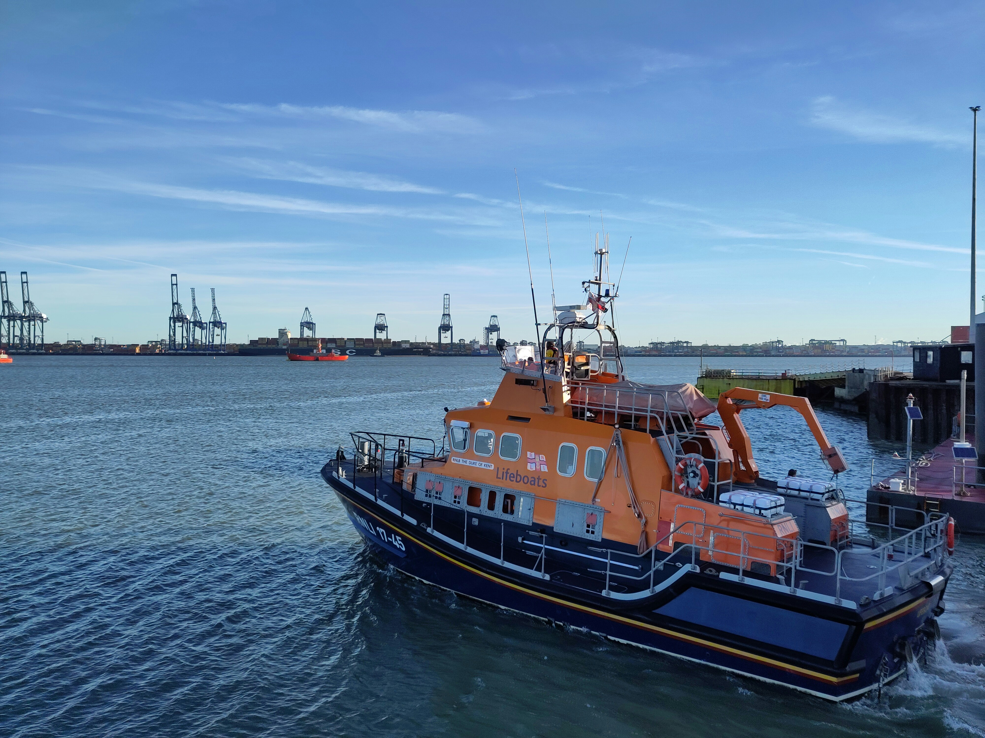 A vibrant orange lifeboat navigates the calm waters of a harbor, with cranes in the background silhouetted against a clear blue sky.