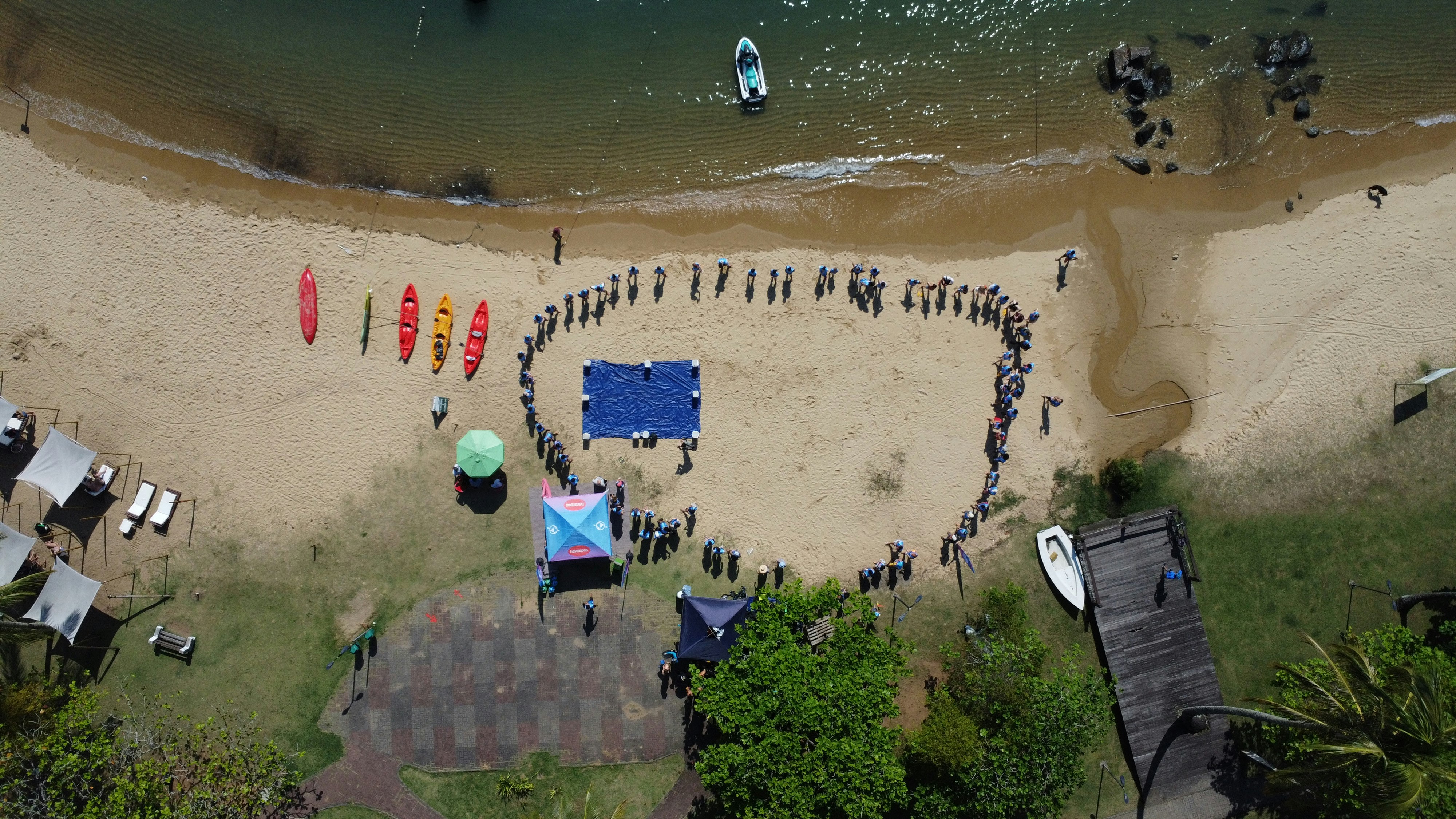 Group forming a circle on a sandy beach with kayaks and trees nearby.