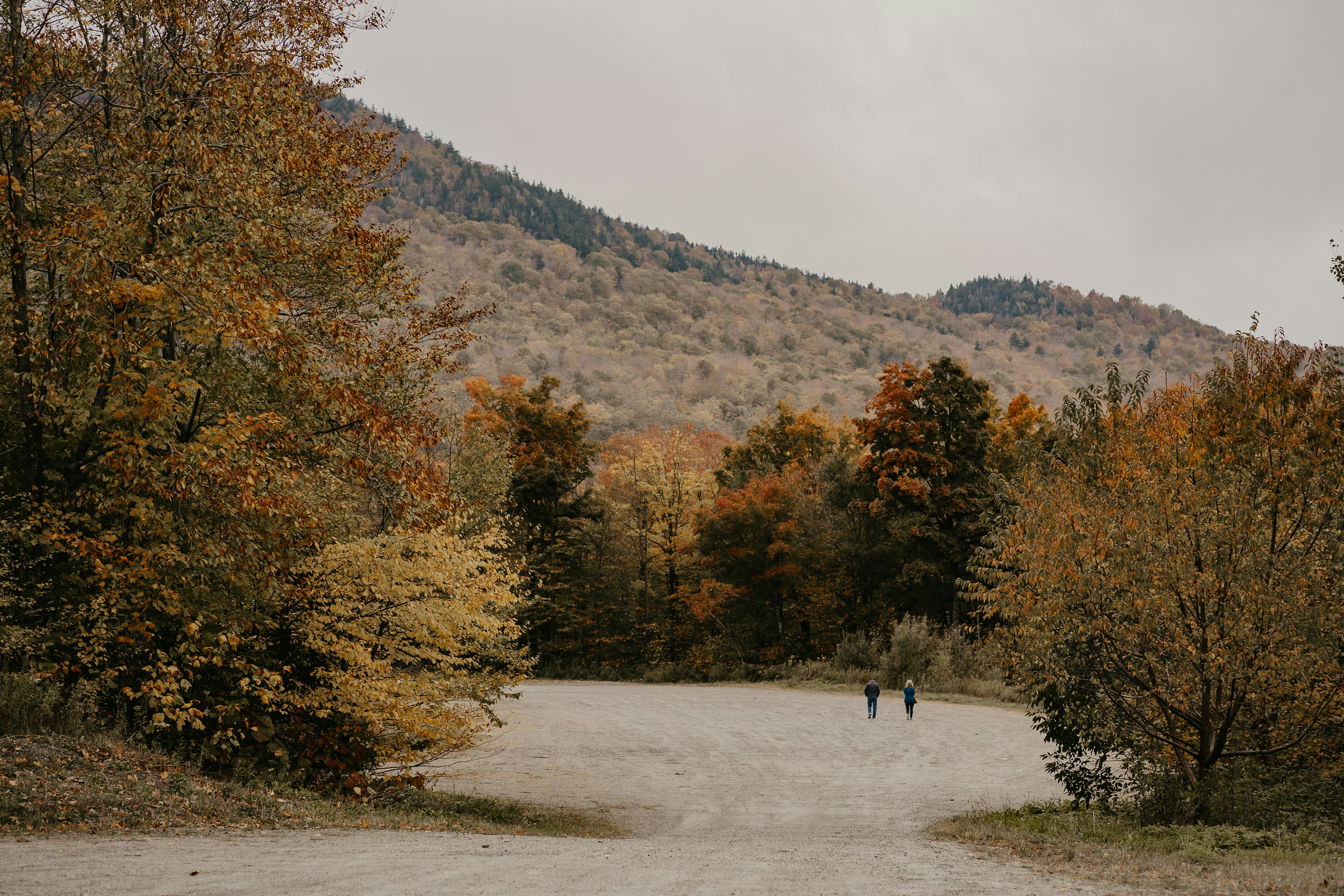 Two people walking on a dirt path surrounded by colorful fall foliage and distant hills.
