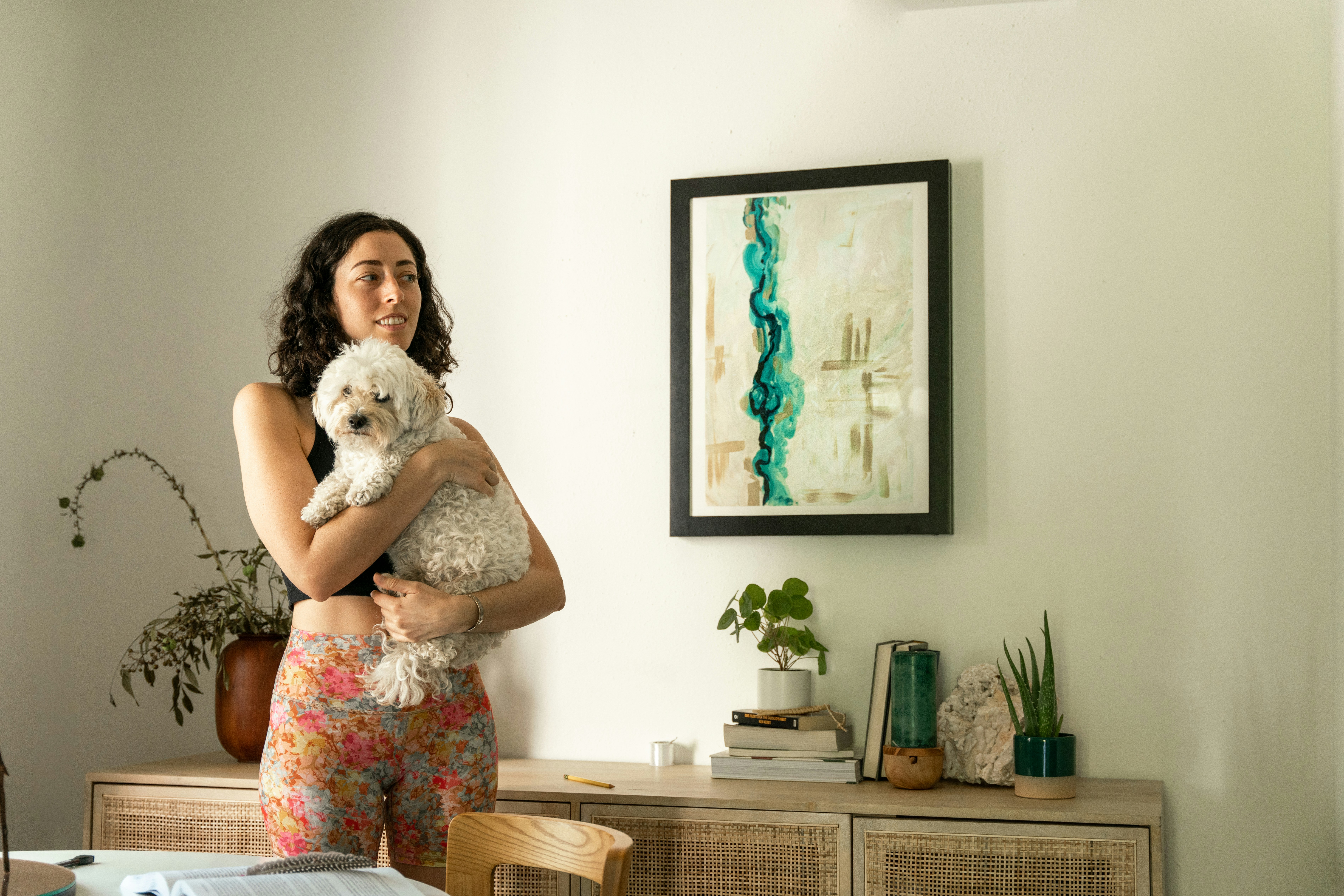 A girl in a tank top carries a white dog in a charming, white and brown interior space. Surrounding them are plants, books, and framed artwork.