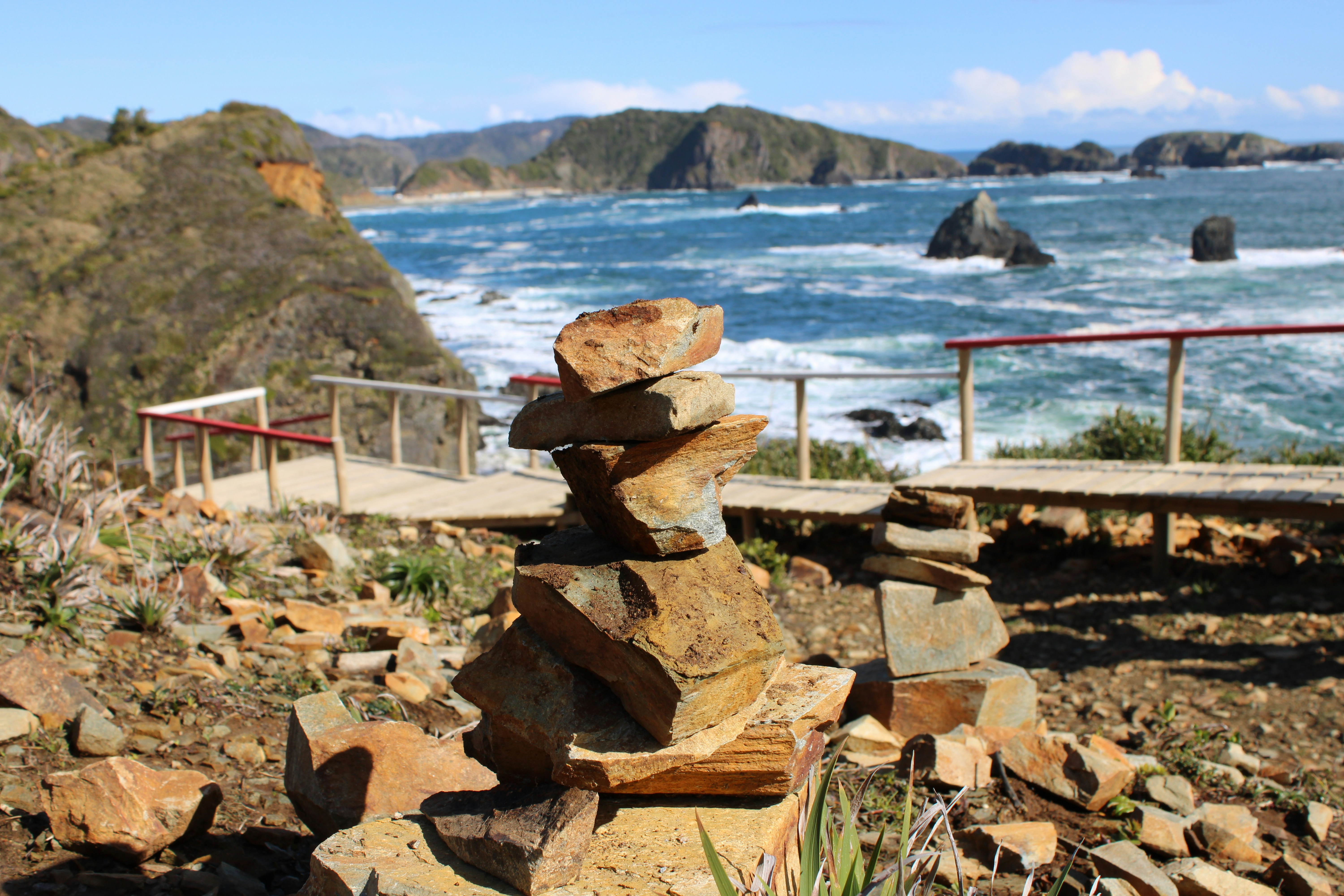 Stacked stones on a coastal path overlook a rugged, wave-swept shoreline under a clear sky.