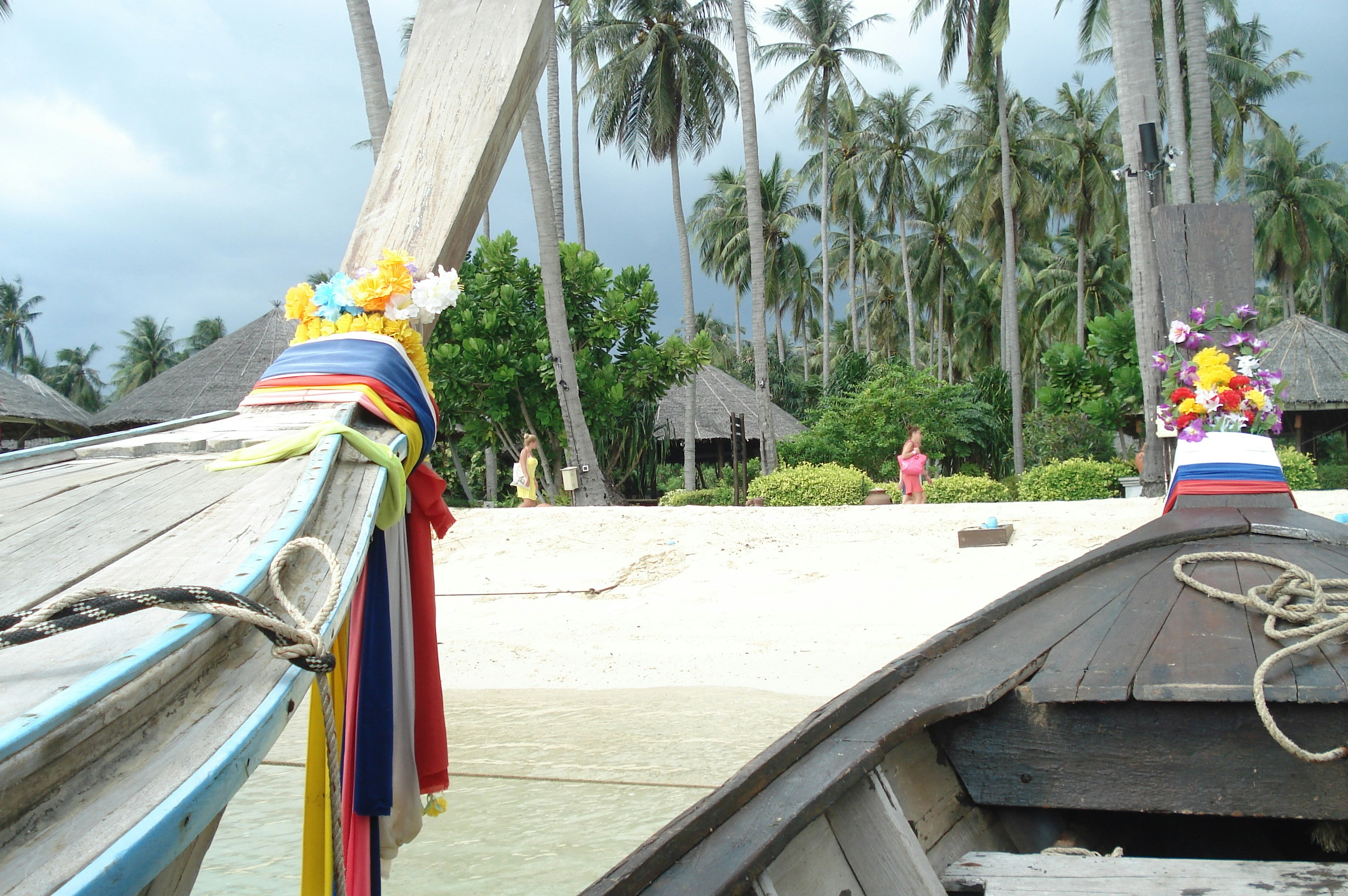 Colorful boat bows adorned with leis and ribbons frame a tropical beach scene with distant figures and tall palm trees.