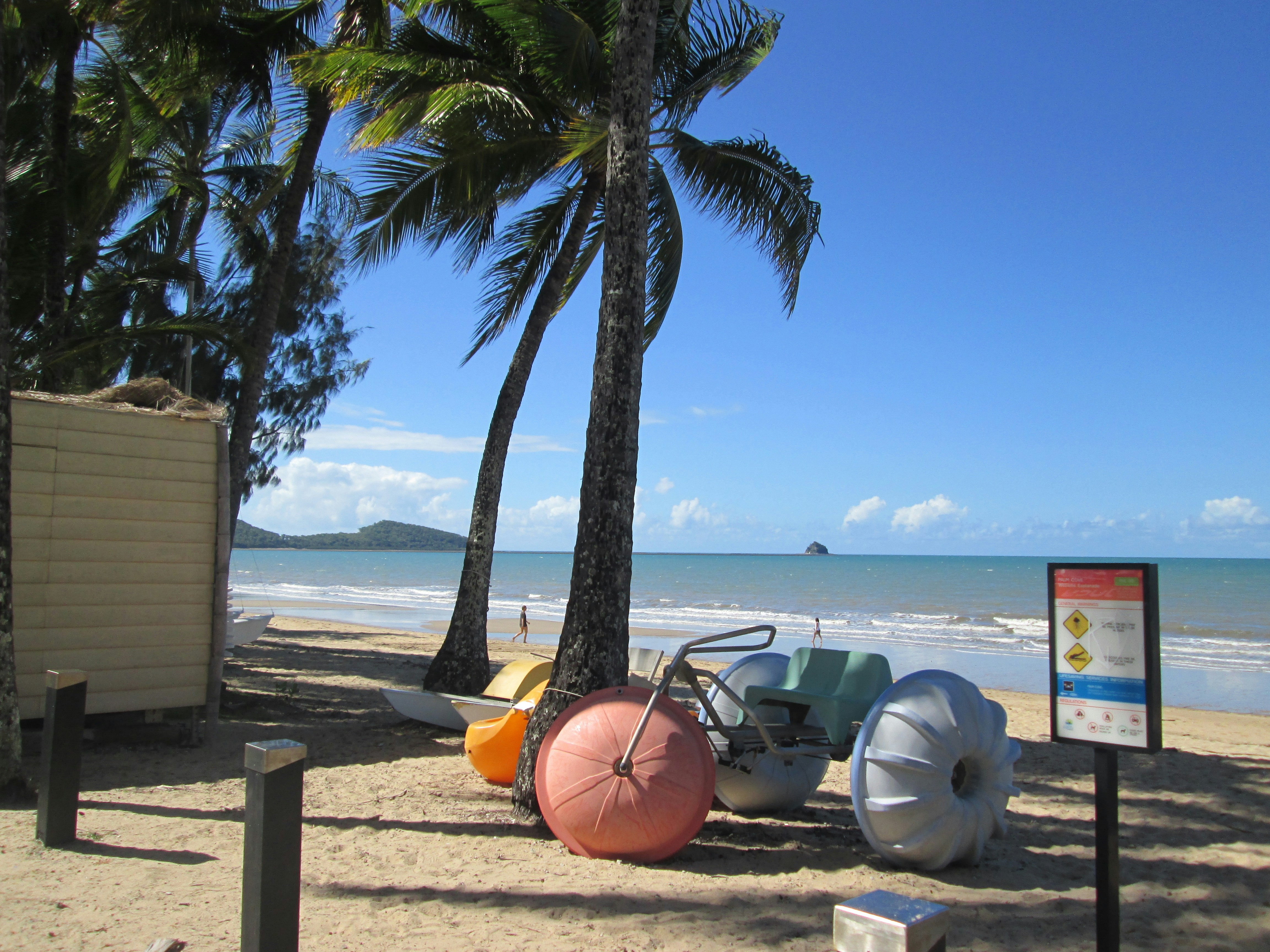 Colorful beach equipment scattered on sandy shore beneath swaying palm trees, with a tranquil sea and distant island in the background.