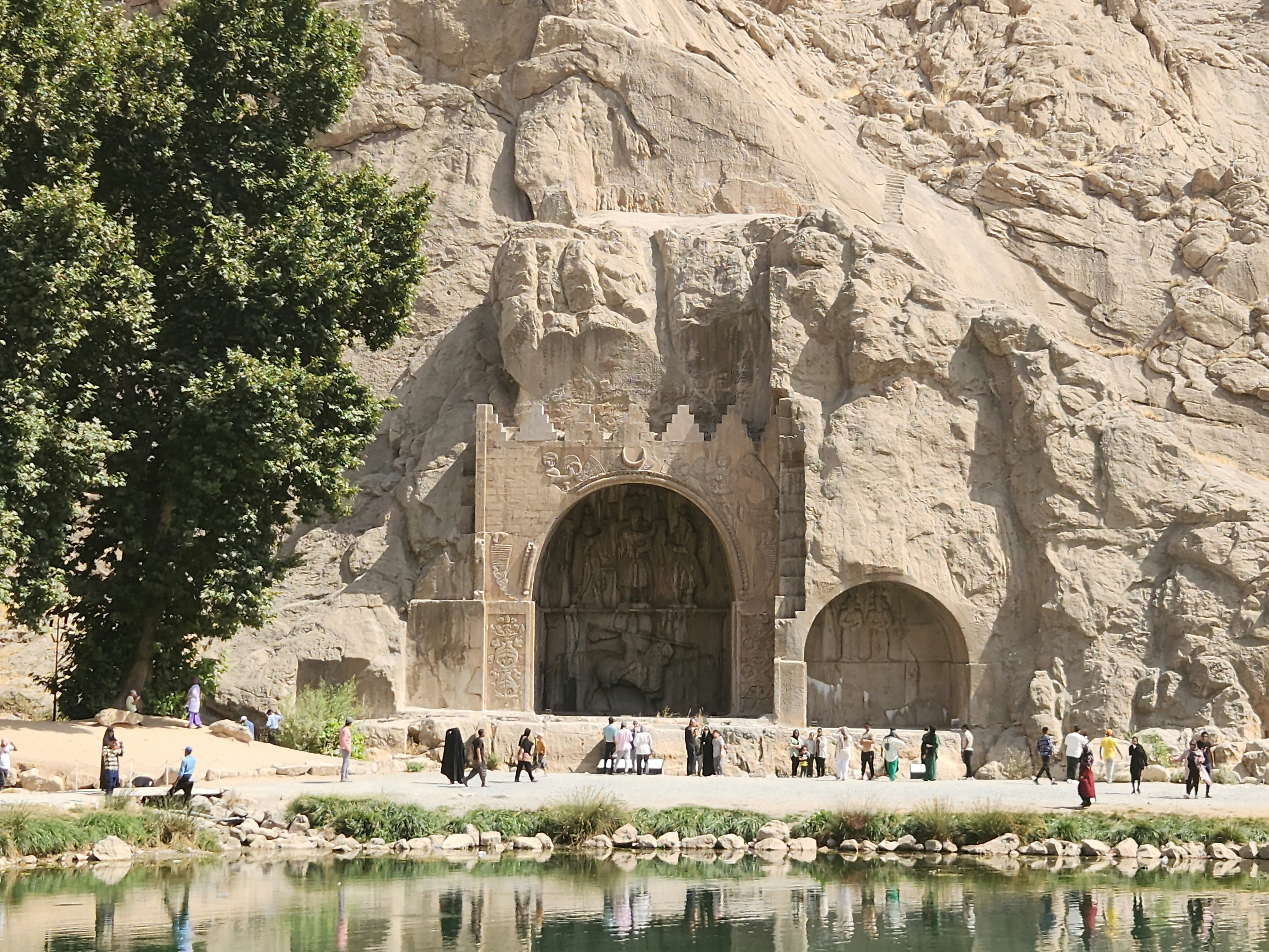 Visitors gather near ancient rock carvings by a tranquil body of water.