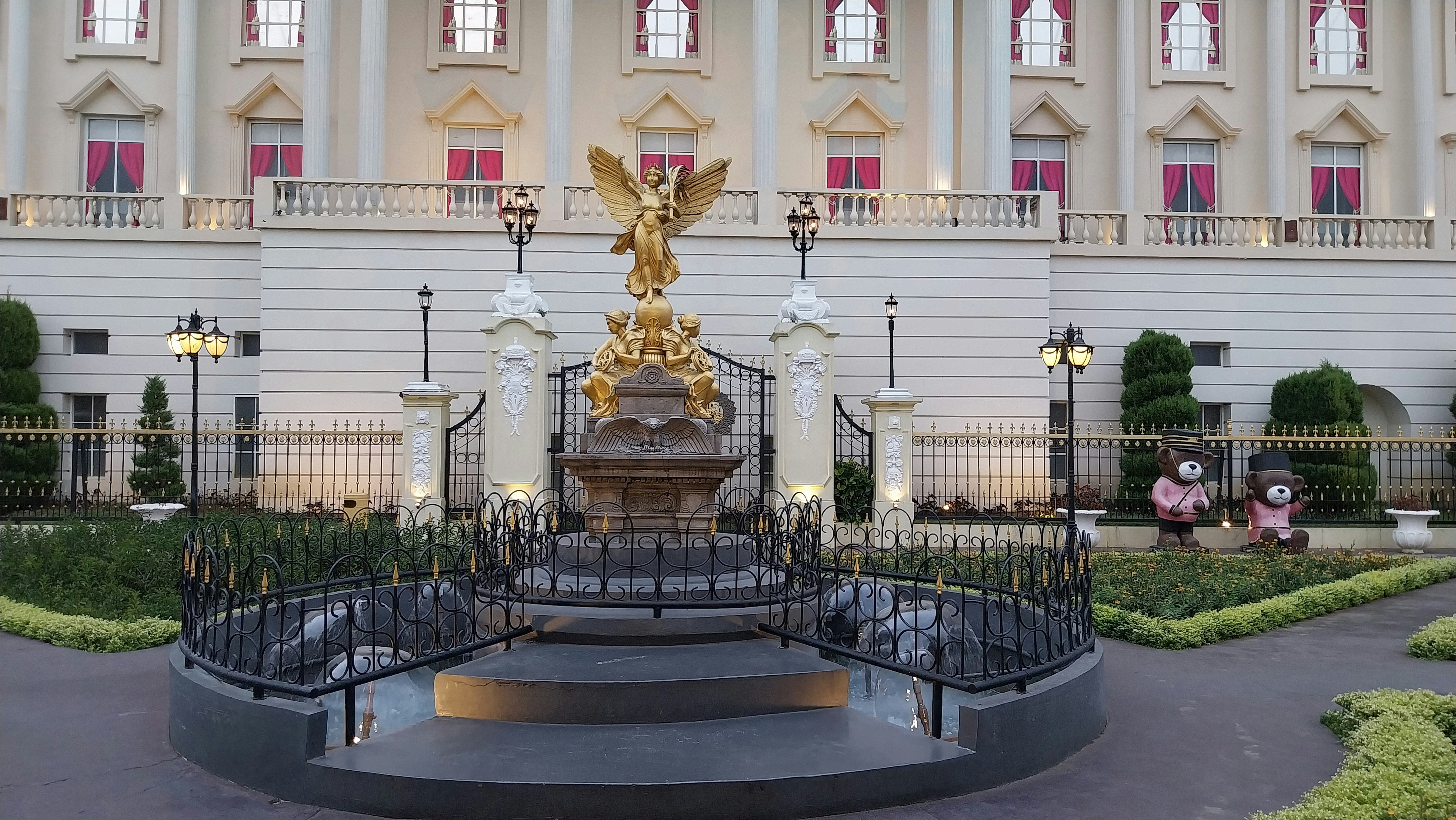 Central gilded statue atop a circular fountain framed by a neoclassical building and manicured gardens. The composition emphasizes symmetry and architectural grandeur.