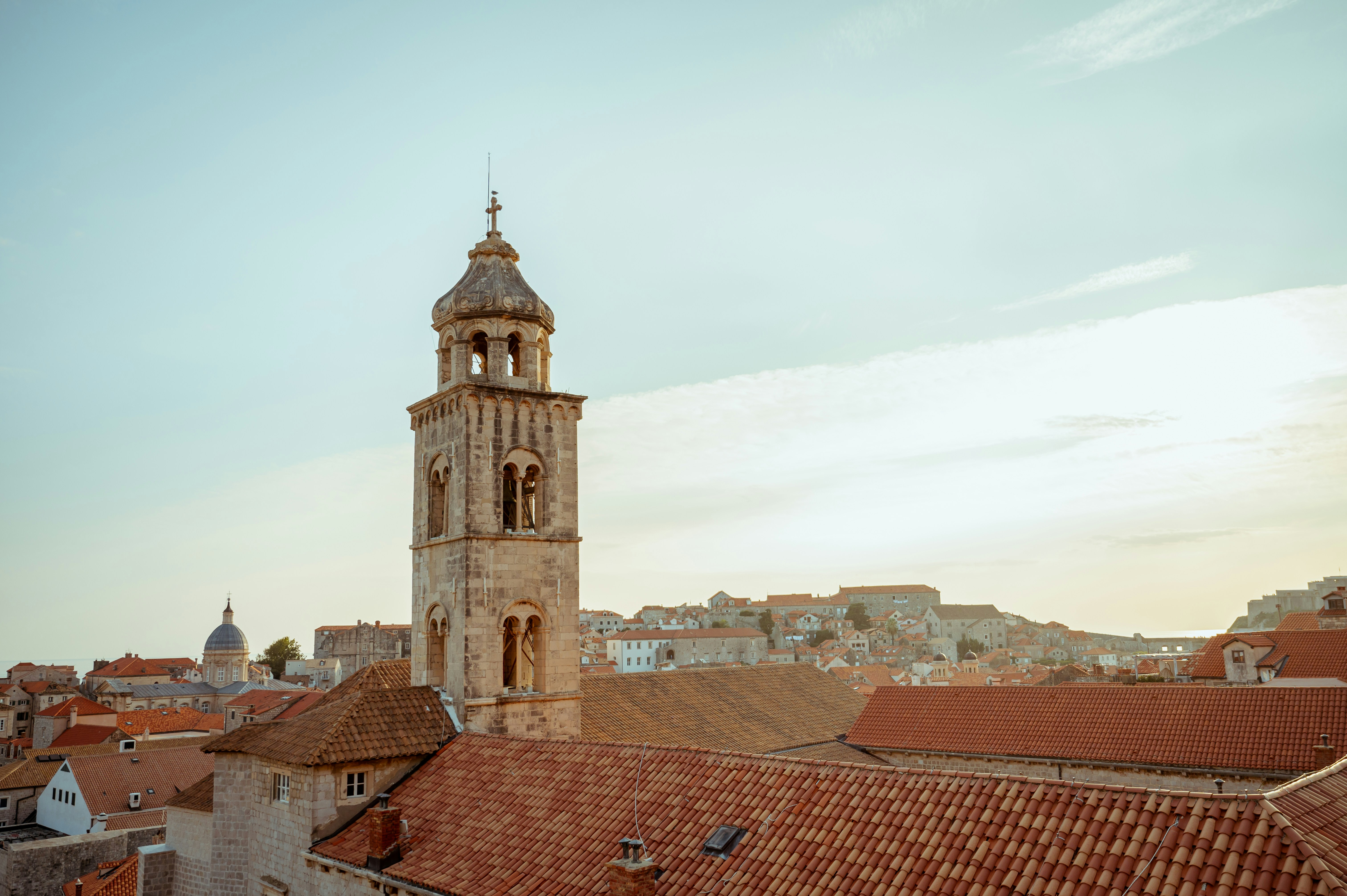 This captivating image captures a picturesque bell tower rising above a sea of terracotta rooftops in a historic European town. The warm glow of the setting sun casts a soft, golden light over the scene, highlighting the textures of the aged stone and clay tiles against a backdrop of a tranquil, pastel sky. The composition draws the eye to the elegant architecture of the tower, creating a harmonious blend of natural and man-made beauty that evokes a sense of timeless serenity.