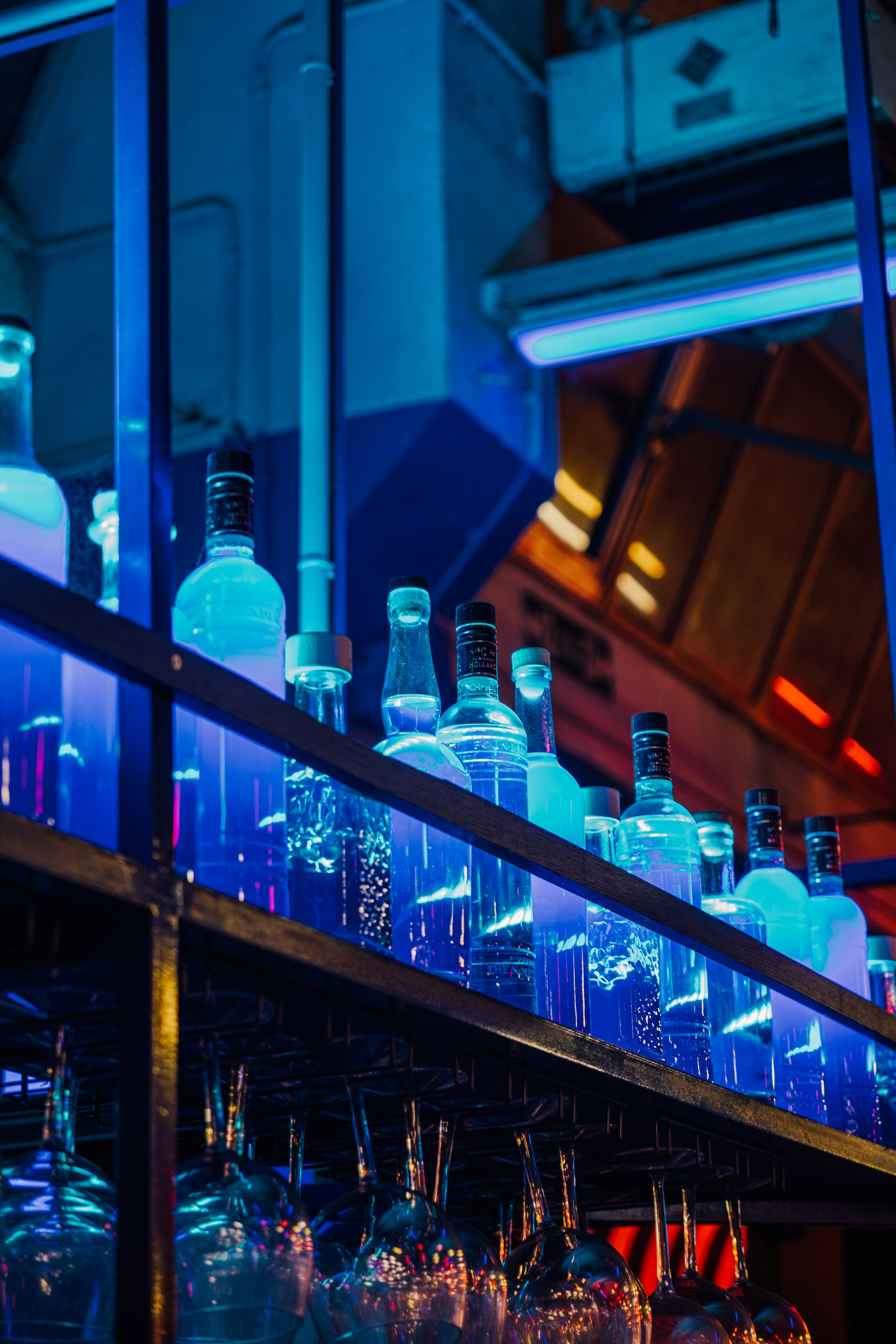 Blue neon-lit bottles line a bar shelf above glassware, bathing the scene in electric blue and purple reflections.