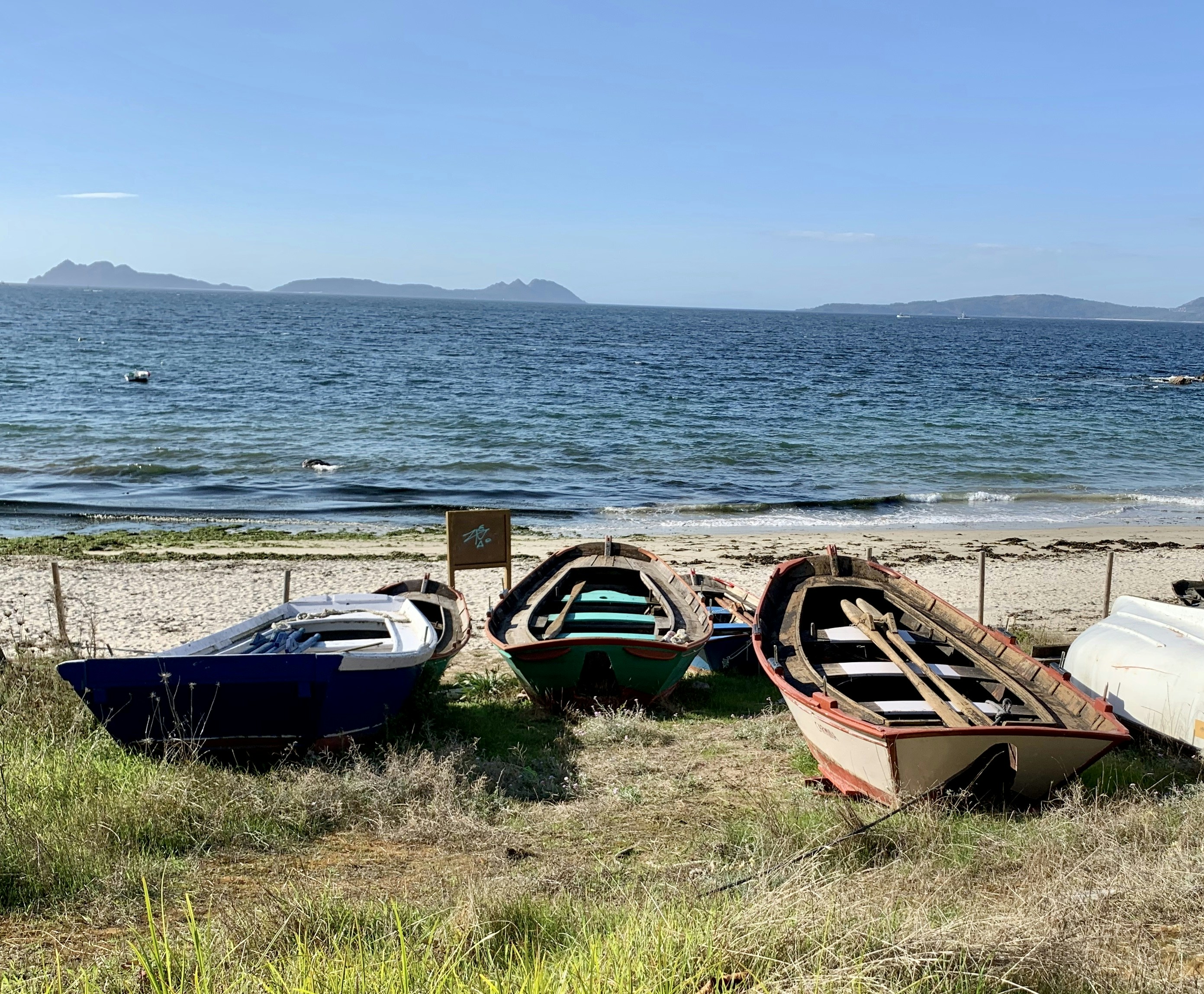 Three colorful boats rest on grassy shore with a calm ocean and distant islands under a clear blue sky.