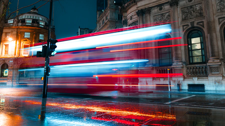 A double-decker bus streaks through a wet downtown street at night, leaving red, white, and blue light trails. Historic stone buildings glow under city lights in the background.