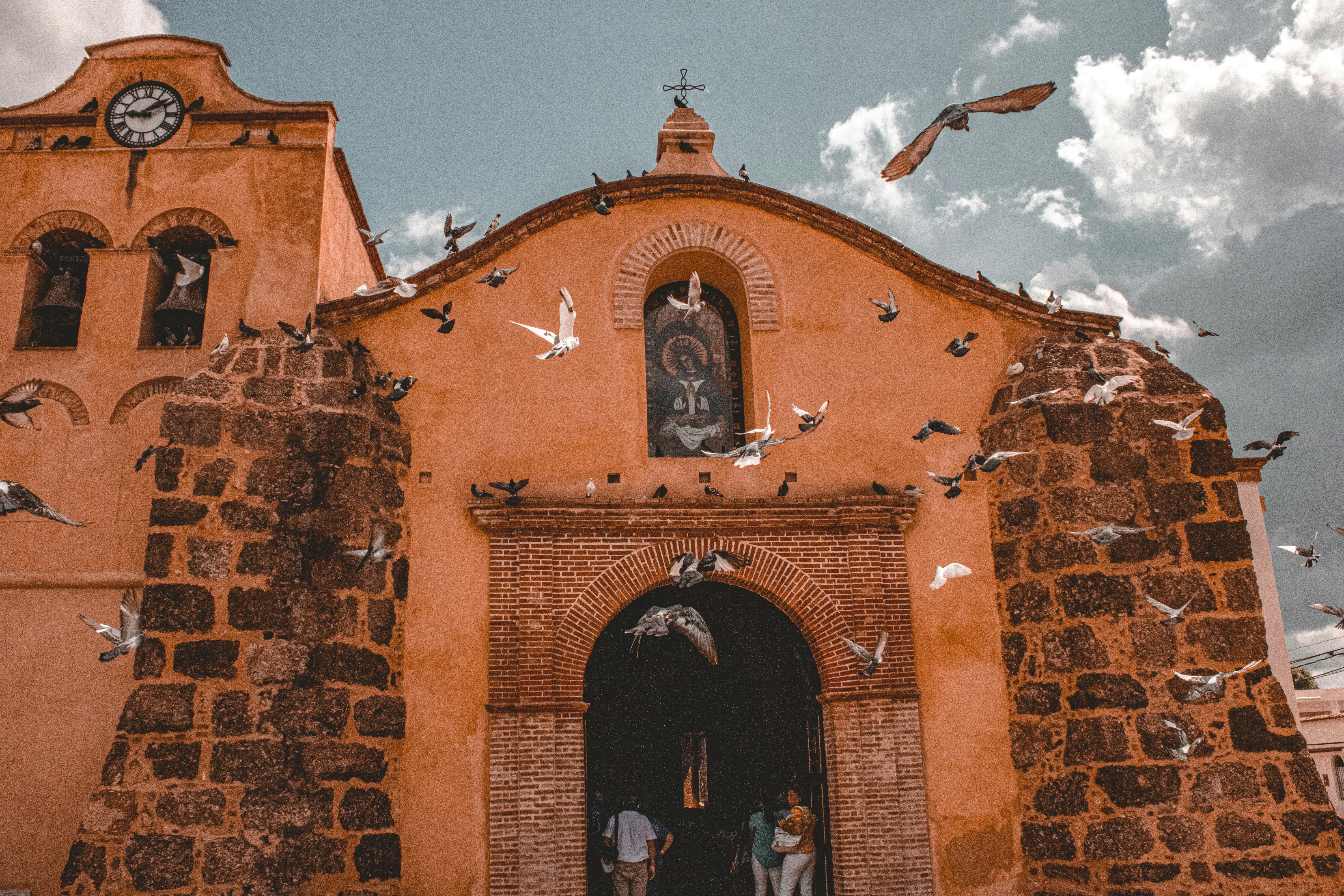 Doves flying around a rustic chapel with a stone façade under a partly cloudy sky.