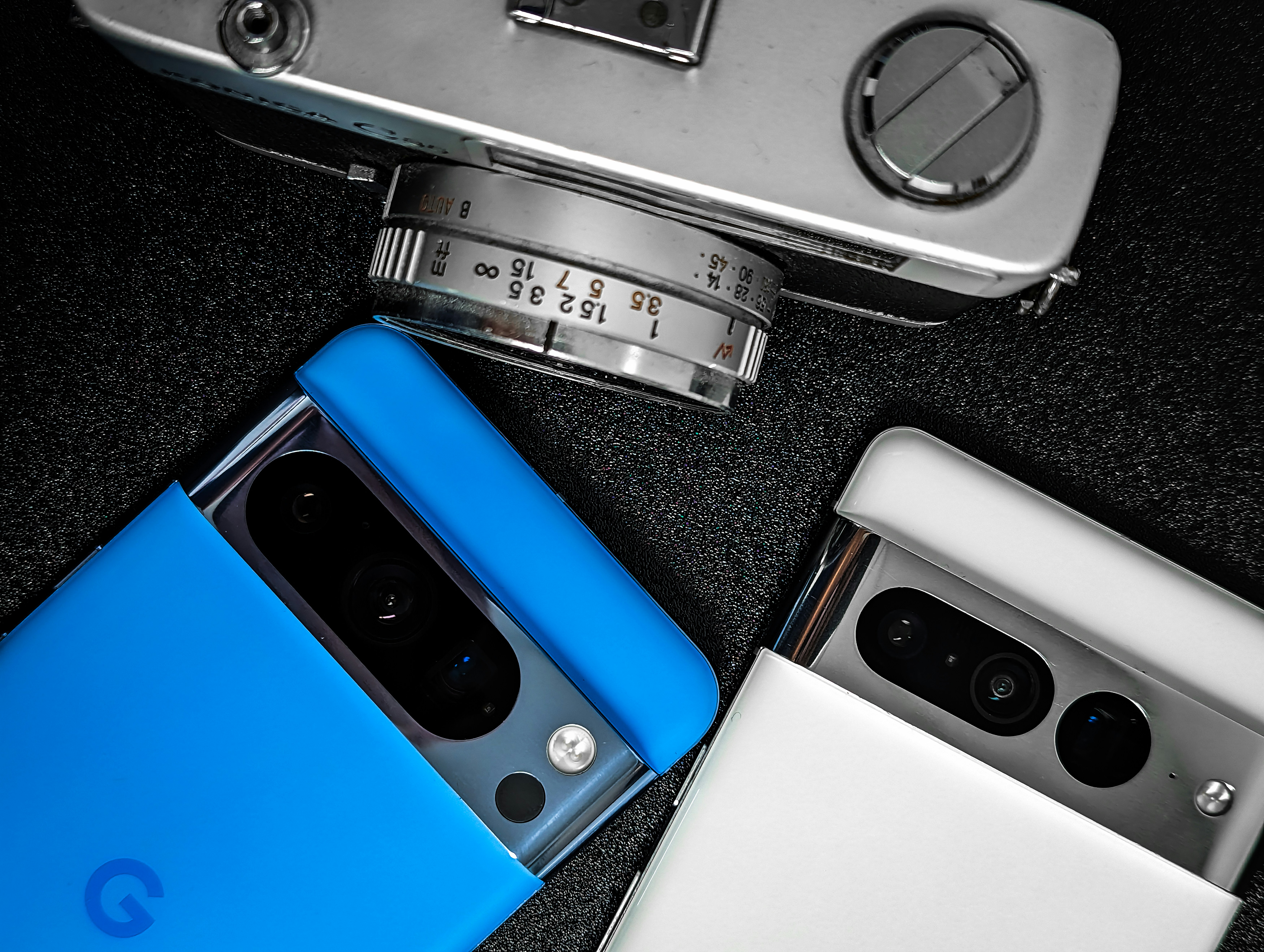 Two Google Pixel 6 Pro phones sit beside a vintage silver camera on a dark textured surface.