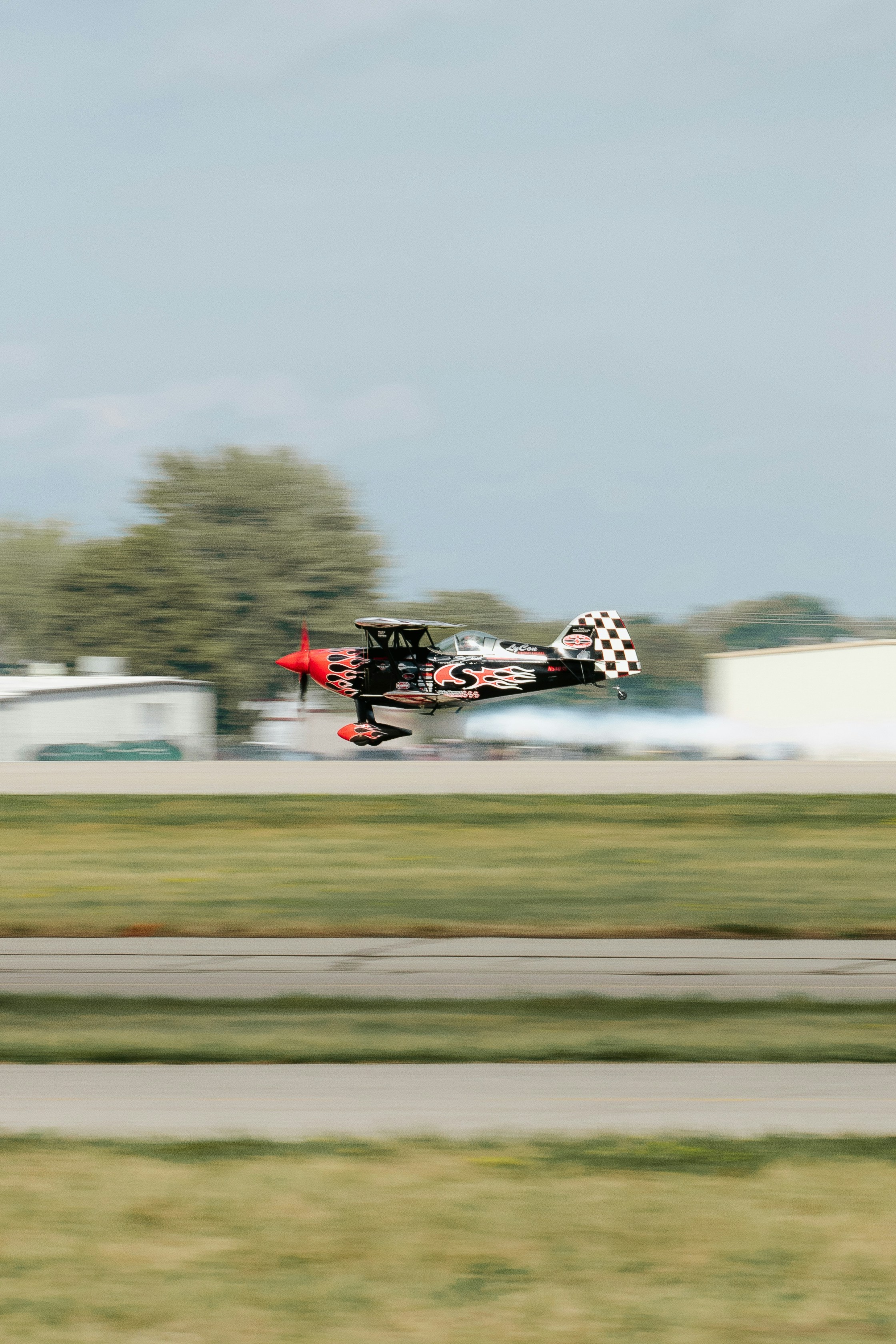 Checkerboard Tailflight: A Red-Nosed Biplane Rises in a Runway Blur