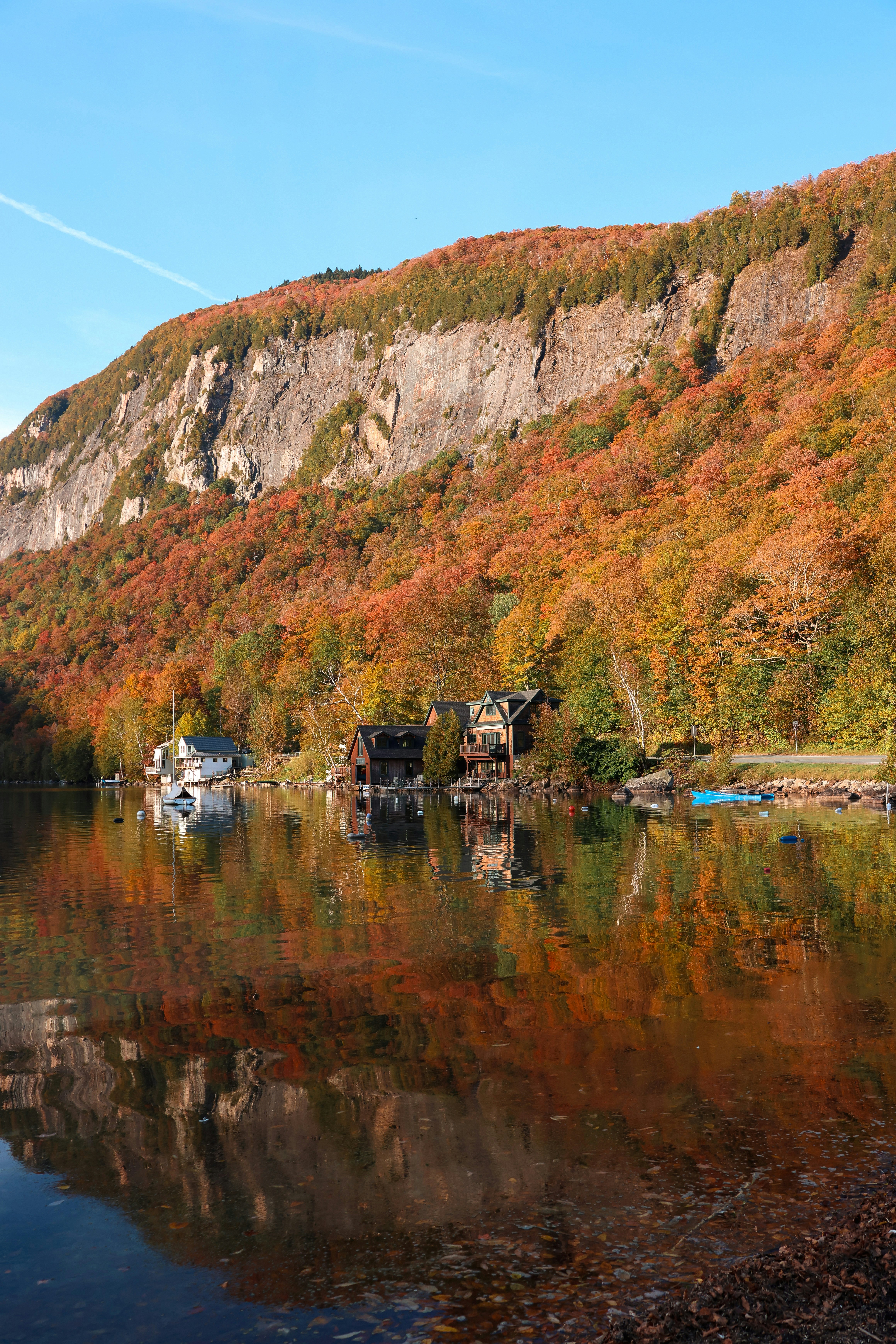 Colorful autumn trees line a tranquil lakeshore with reflected houses and boats under a steep hillside. A calm scene of seasonal change by the water.