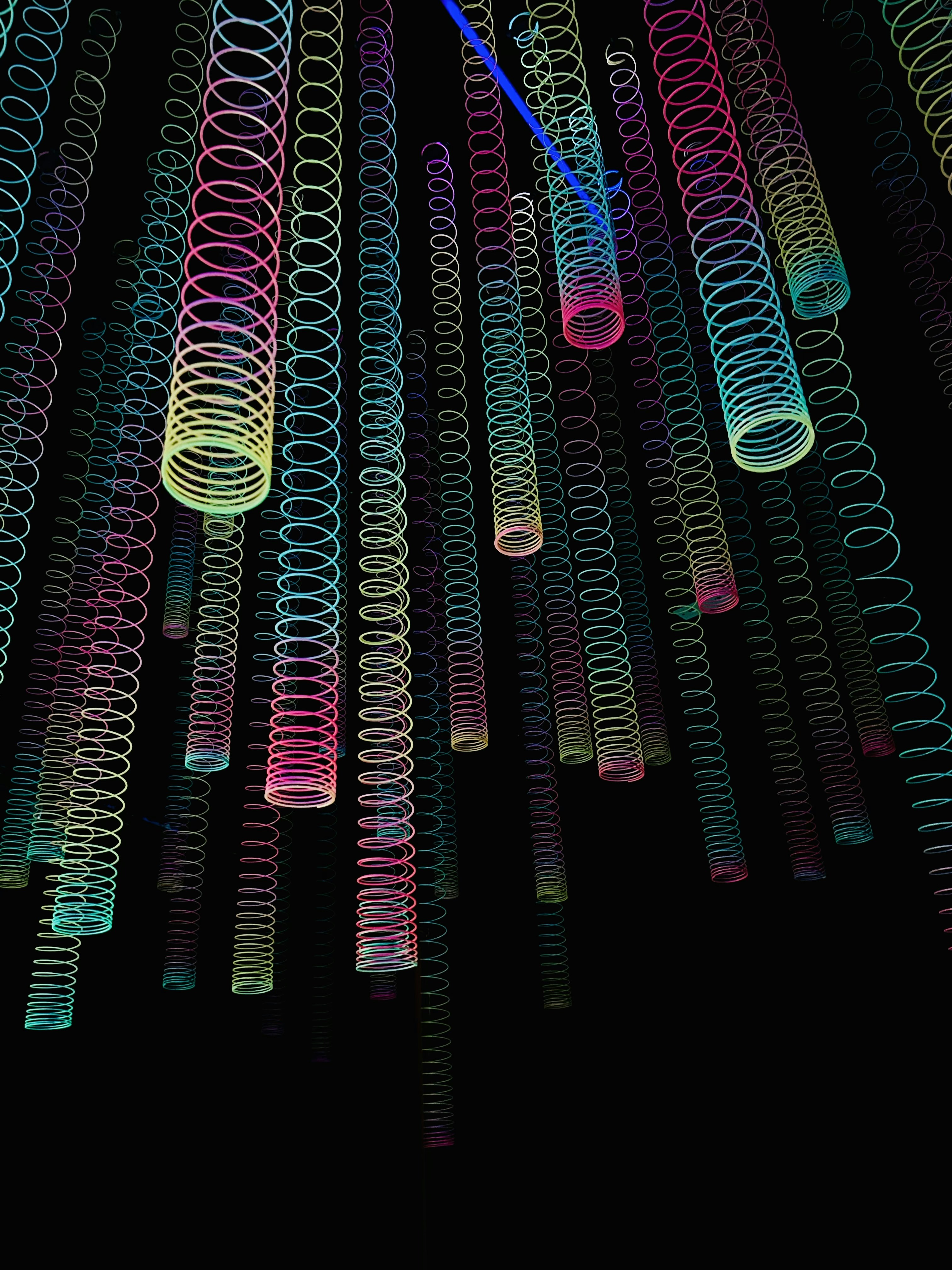 Vertical cascade of multicolored neon coils suspended against a black background. This digital art installation emphasizes color, rhythm, and depth.