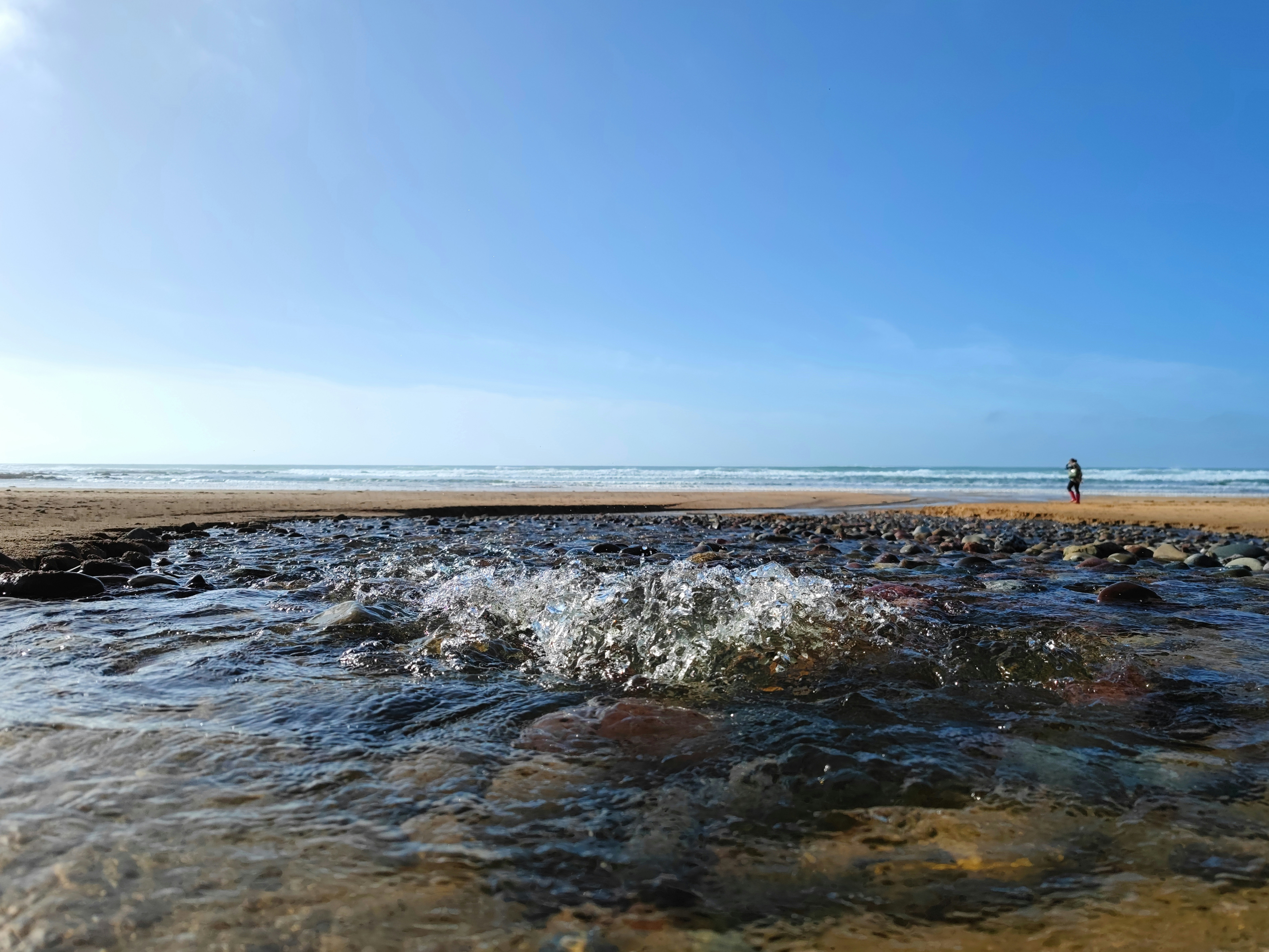 Foaming water spills over dark, slick rocks in the foreground as a solitary figure crosses the sunlit beach beneath a clear blue sky.