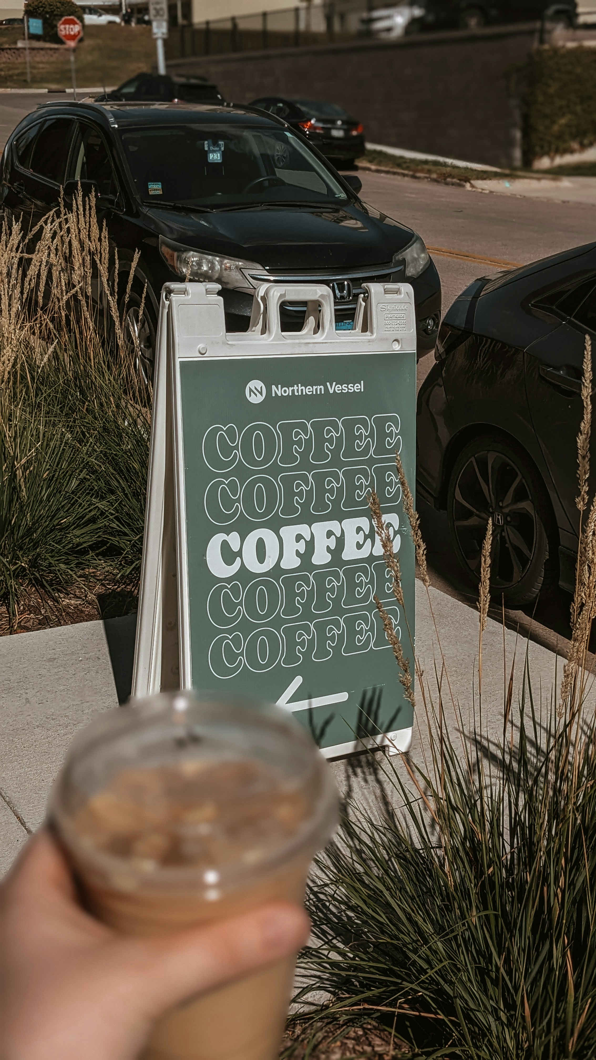Sidewalk coffee signboard in front of parked cars, with a hand holding an iced coffee in the foreground.