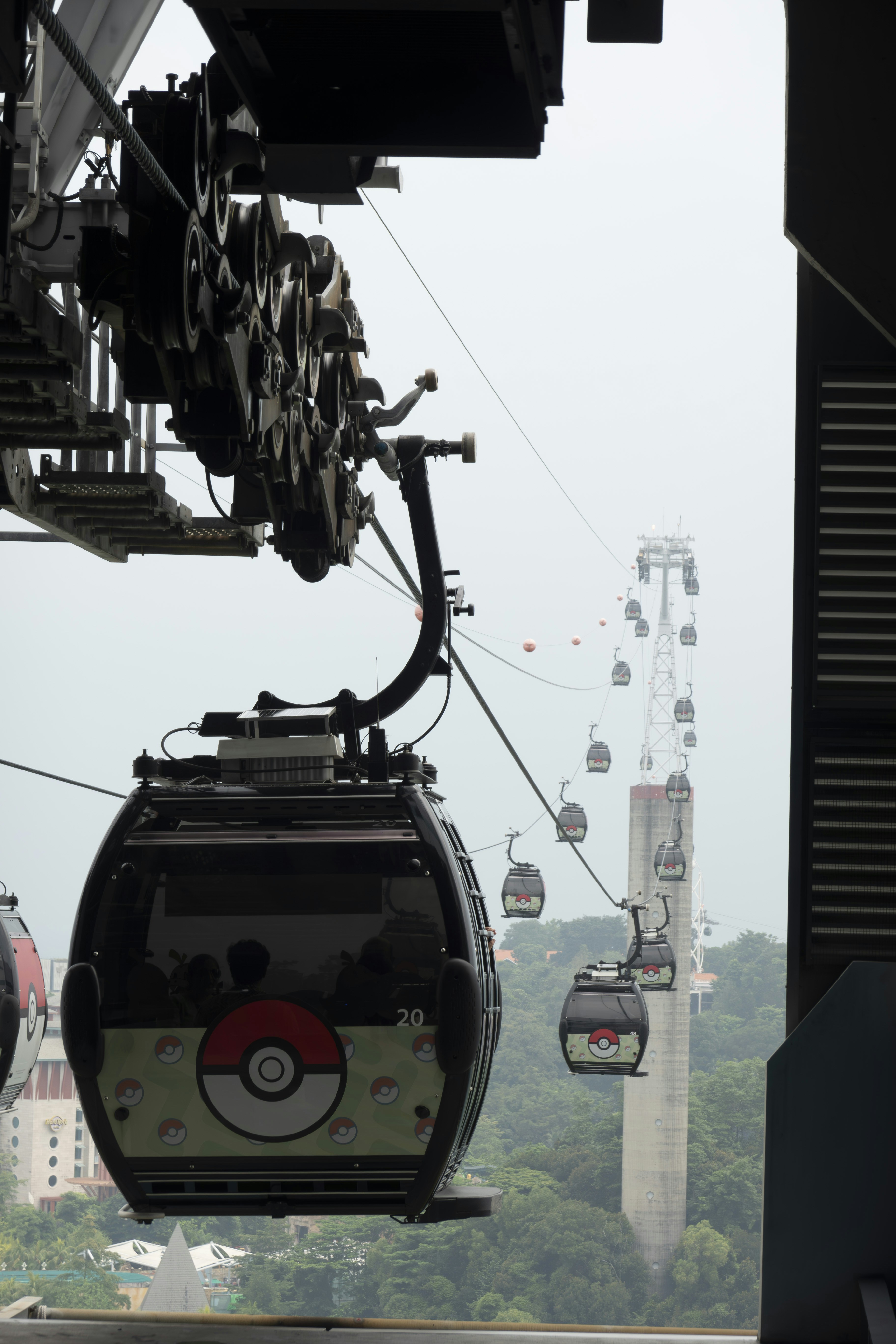Cable car gondola with Poké Ball logo hangs from a cable system, with distant gondolas and a tall tower visible in the background.