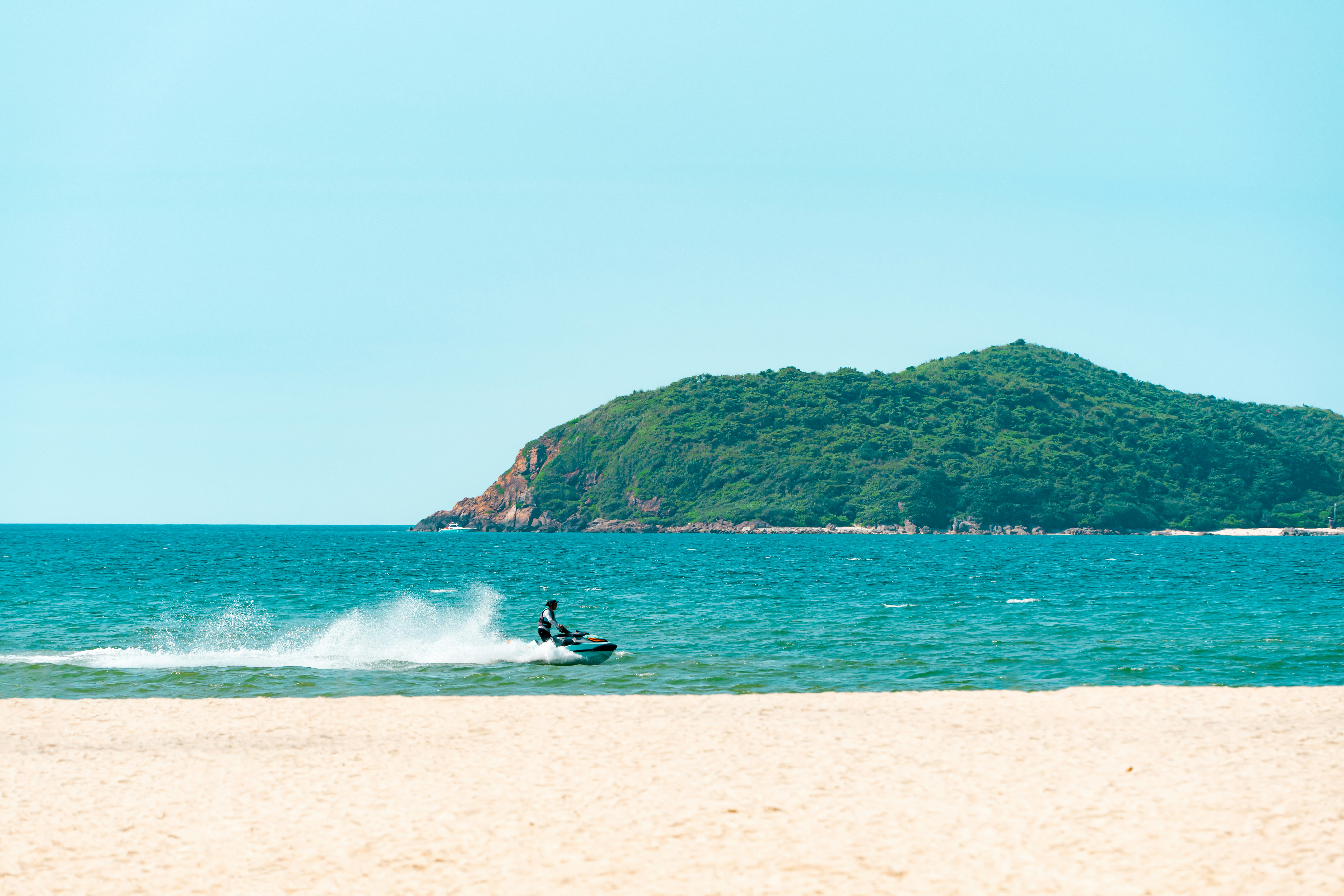 A jet skier glides across the vibrant blue ocean waters, with a lush green island in the background. The sandy beach frames the scene perfectly.
