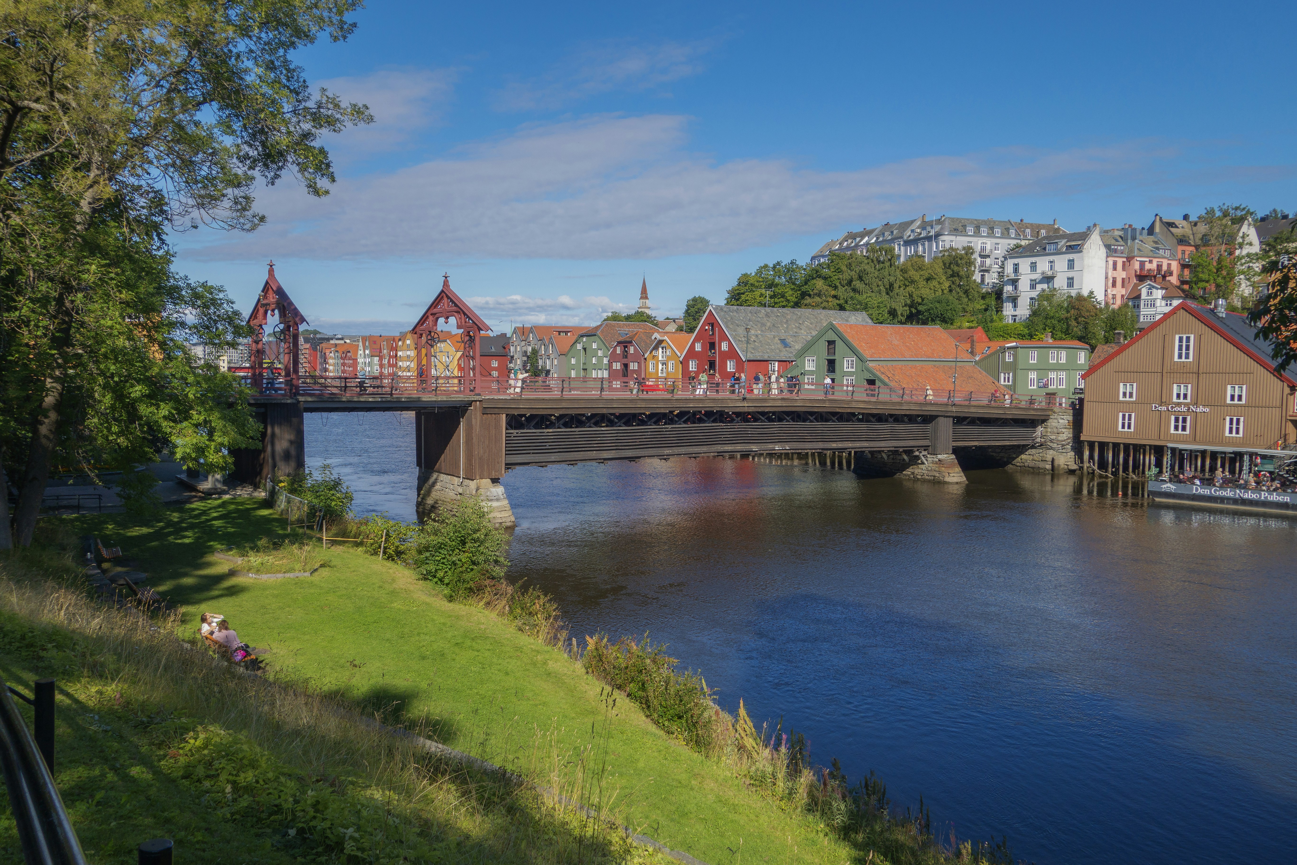 Colorful wooden buildings line a bridge over a calm river, surrounded by lush greenery under a clear blue sky.