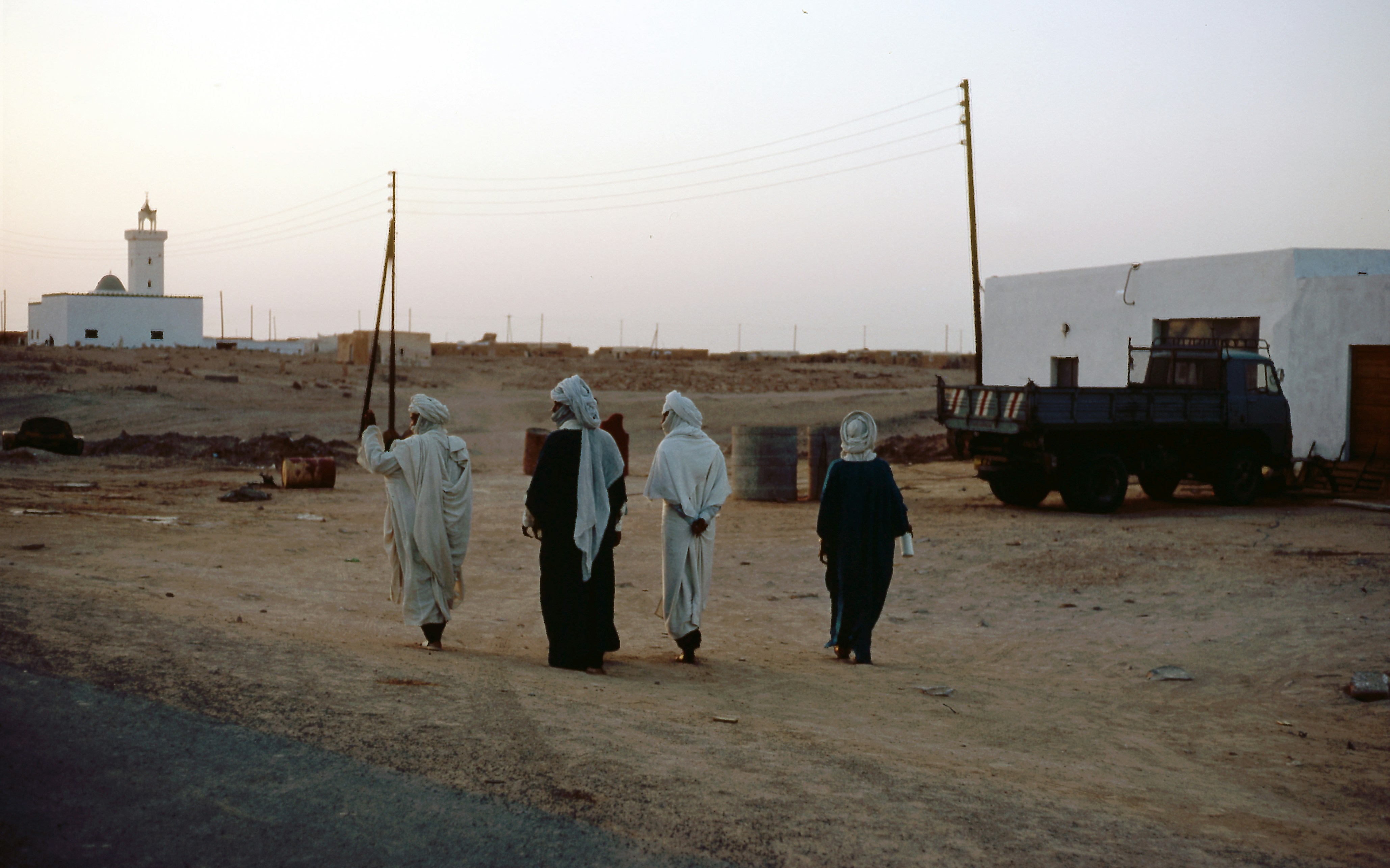 Four figures in traditional attire walk along a dirt road in a desert settlement with distant structures.