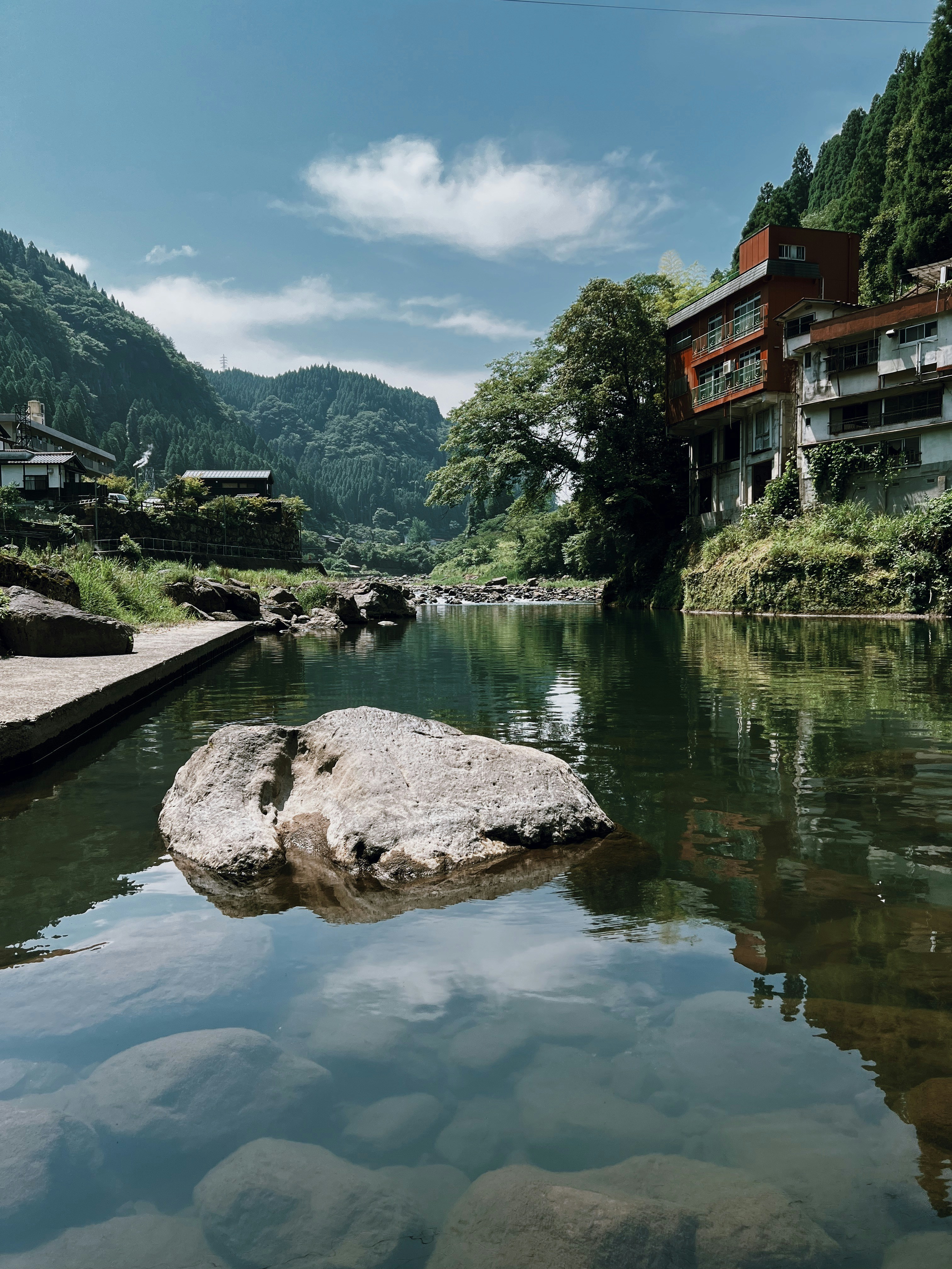 Calm river reflecting surrounding greenery and a rustic building under a clear blue sky.