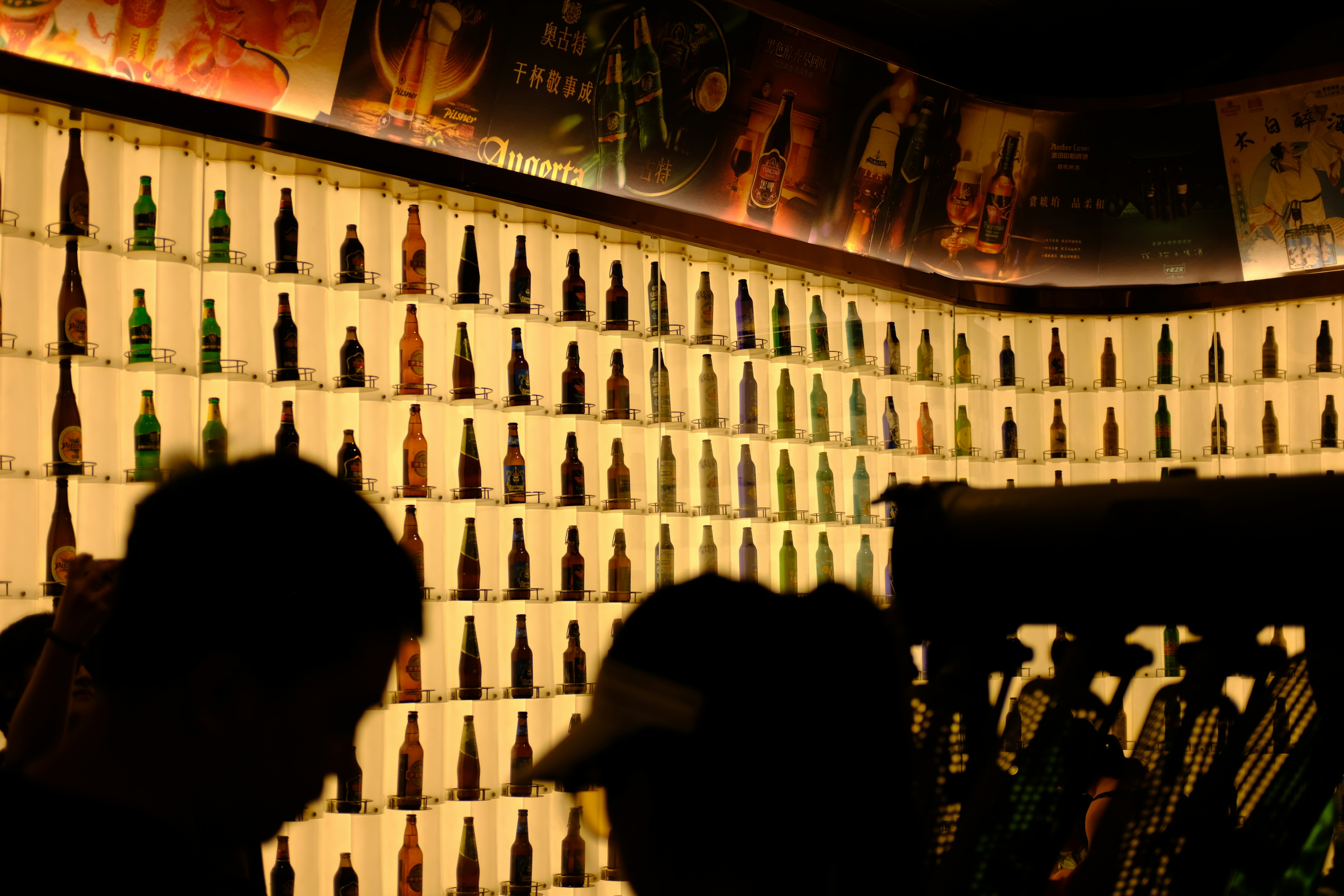 Silhouetted patrons against a backlit wall of neatly arranged beer bottles in a dimly lit bar.