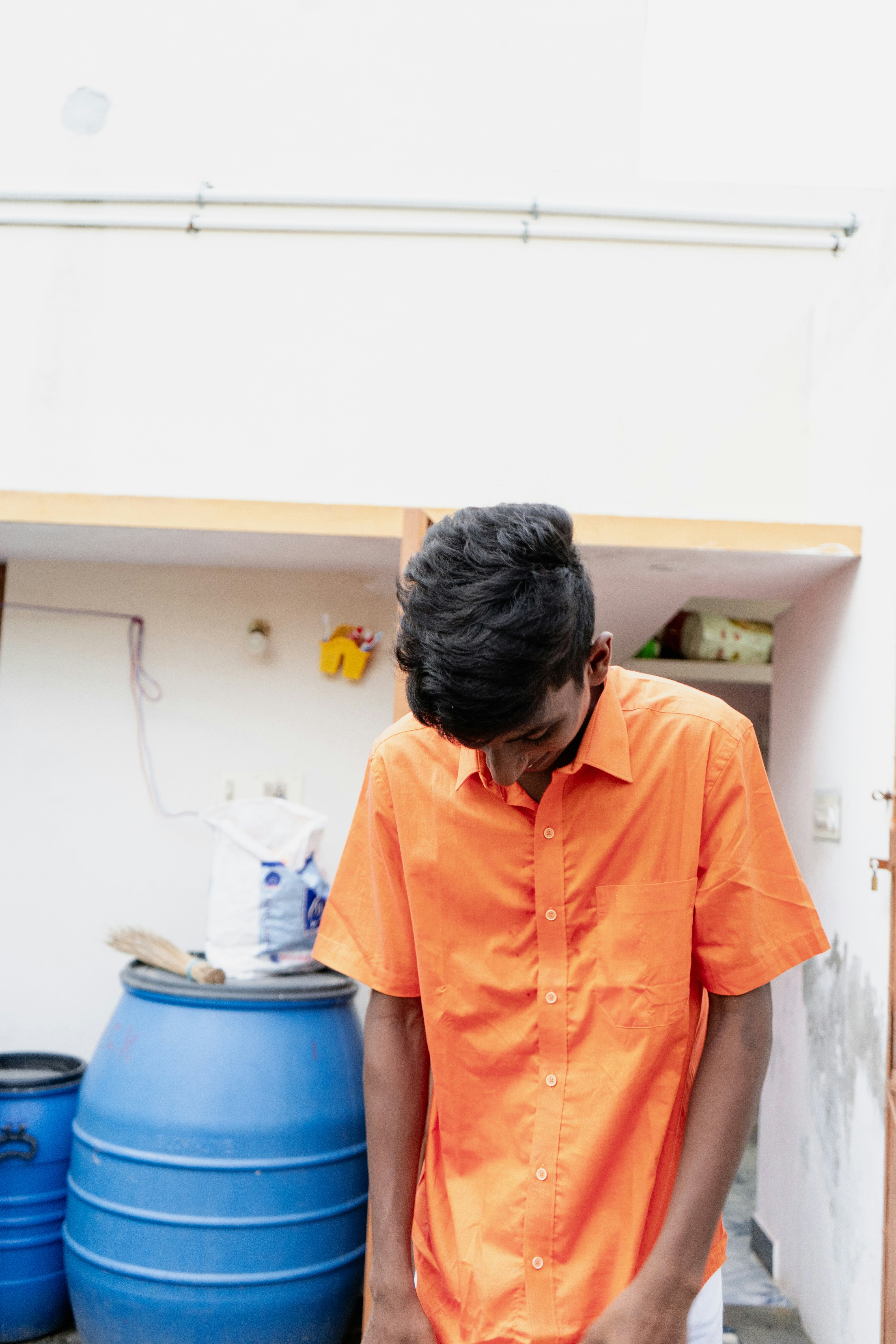 Contemplative Moment in a Sunlit Utility Corner: Orange Shirt, Blue Barrels