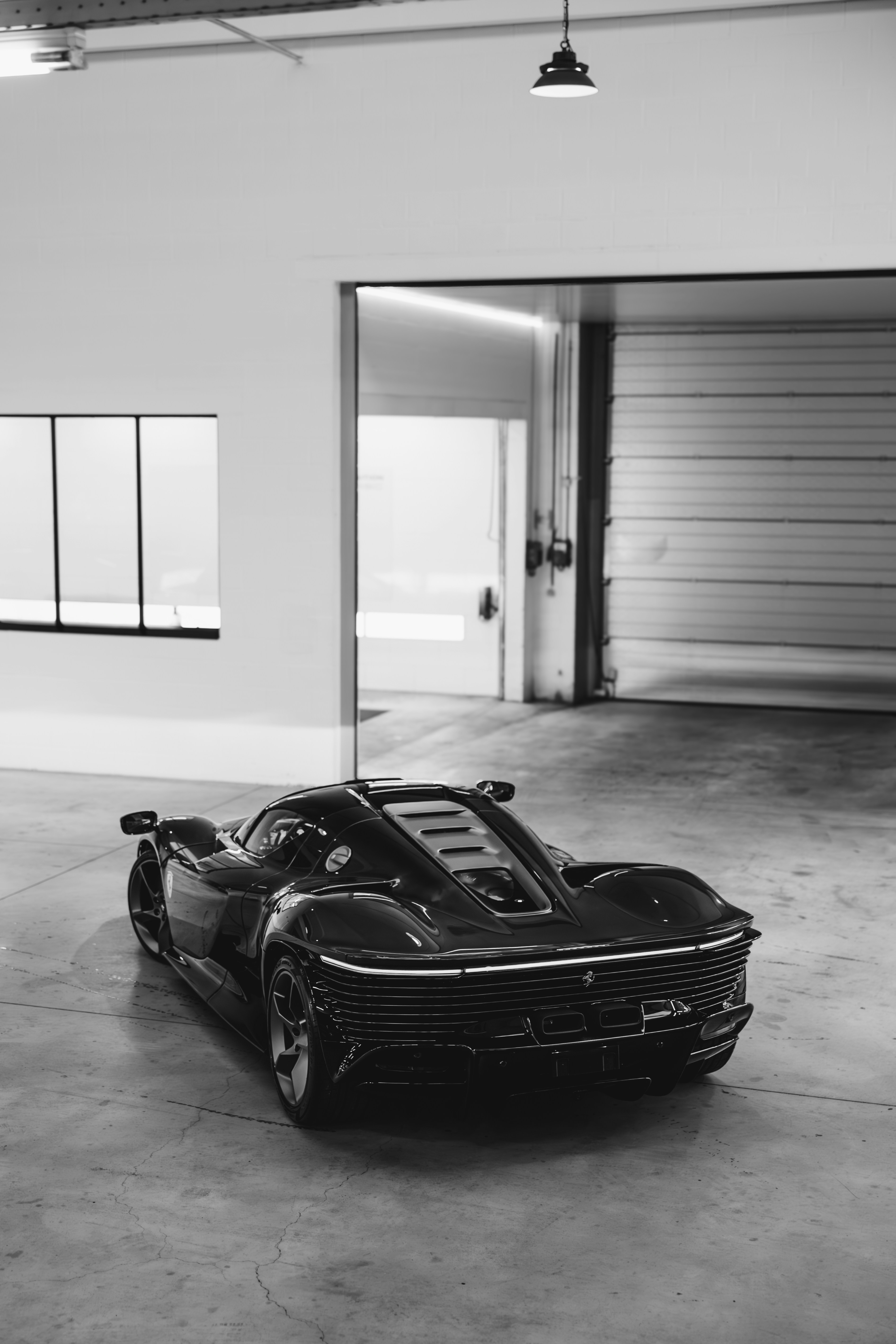 Monochrome photograph of a sleek black hypercar parked inside a bright, empty warehouse with a rolling shutter in the background.
