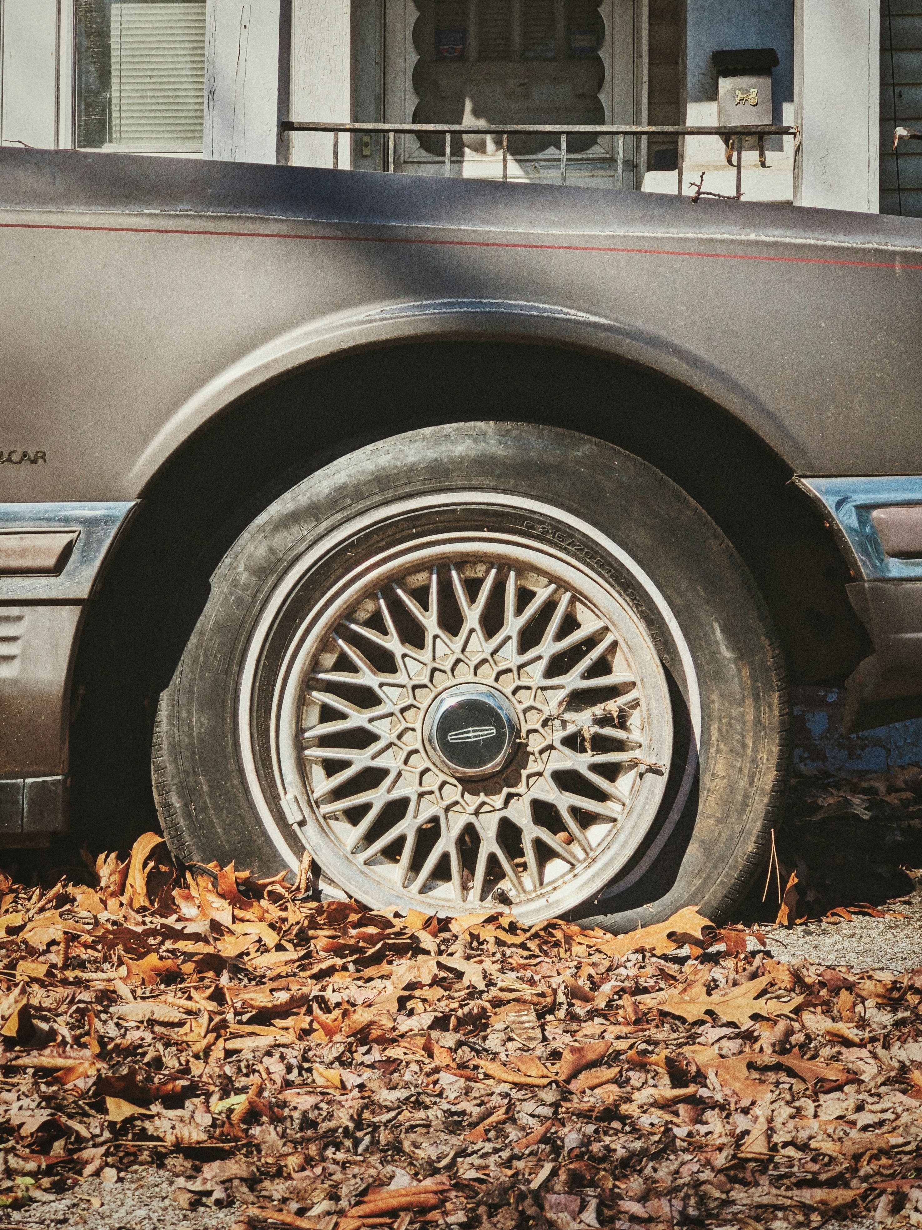 The front wheel of an abandoned Cadillac Town Car with fallen leaves scattered in front of it.