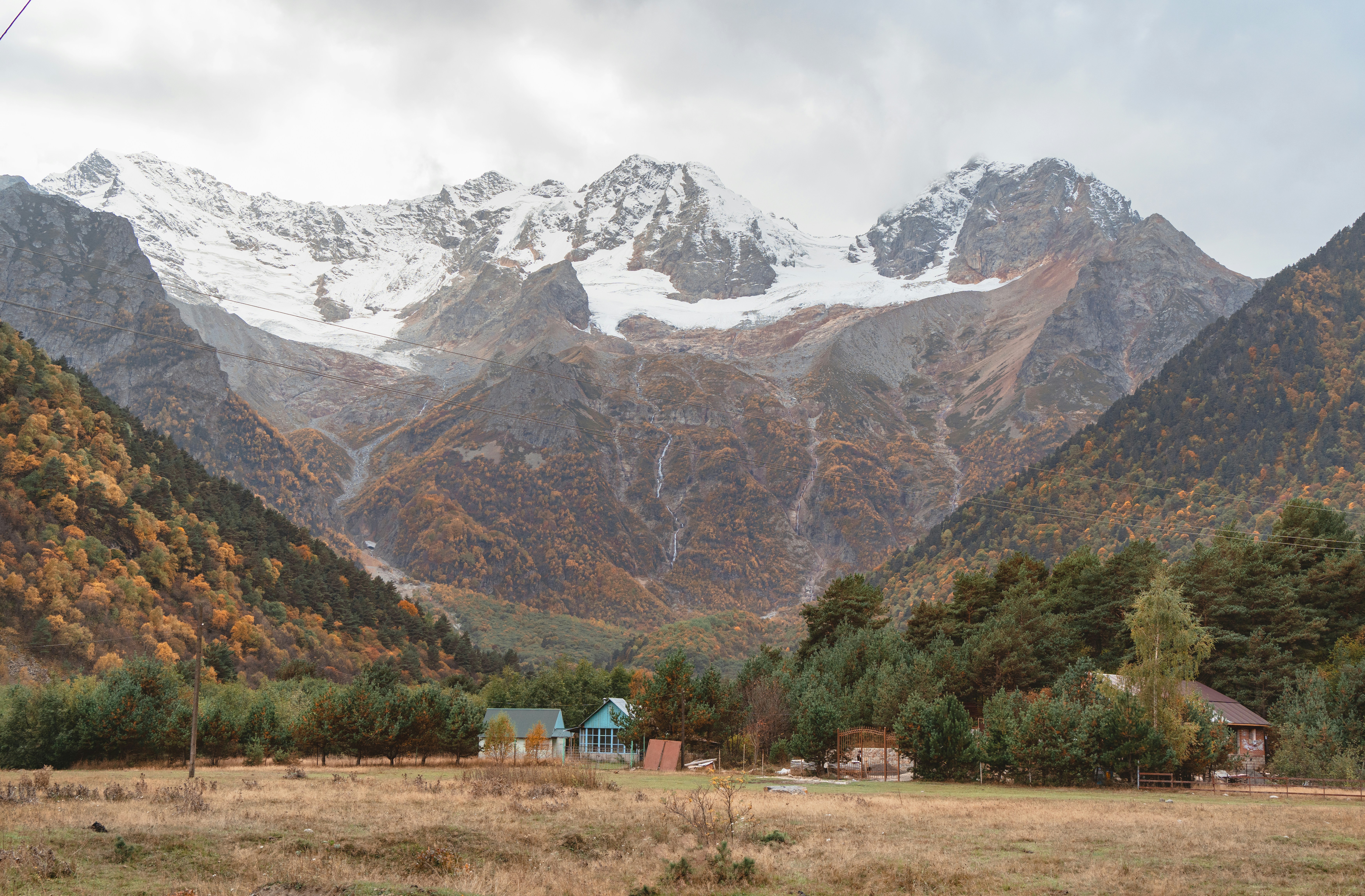 Mountain range with snow-capped peaks and a rustic house in the foreground, framed by autumn foliage.
