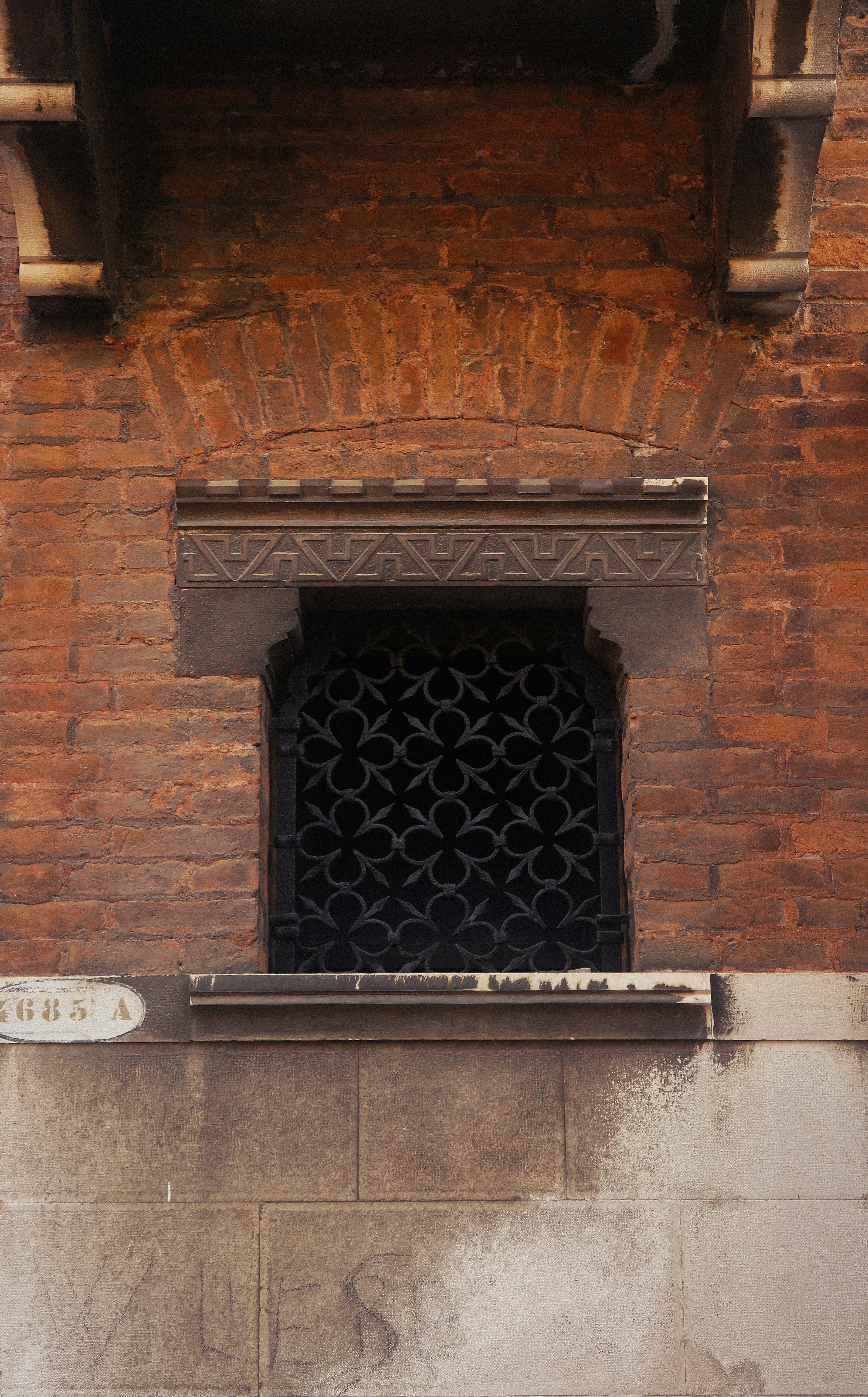 Central brick façade frames a small arched opening protected by a geometric iron grille. The composition emphasizes weathered texture and architectural detail bathed in warm, subdued light.