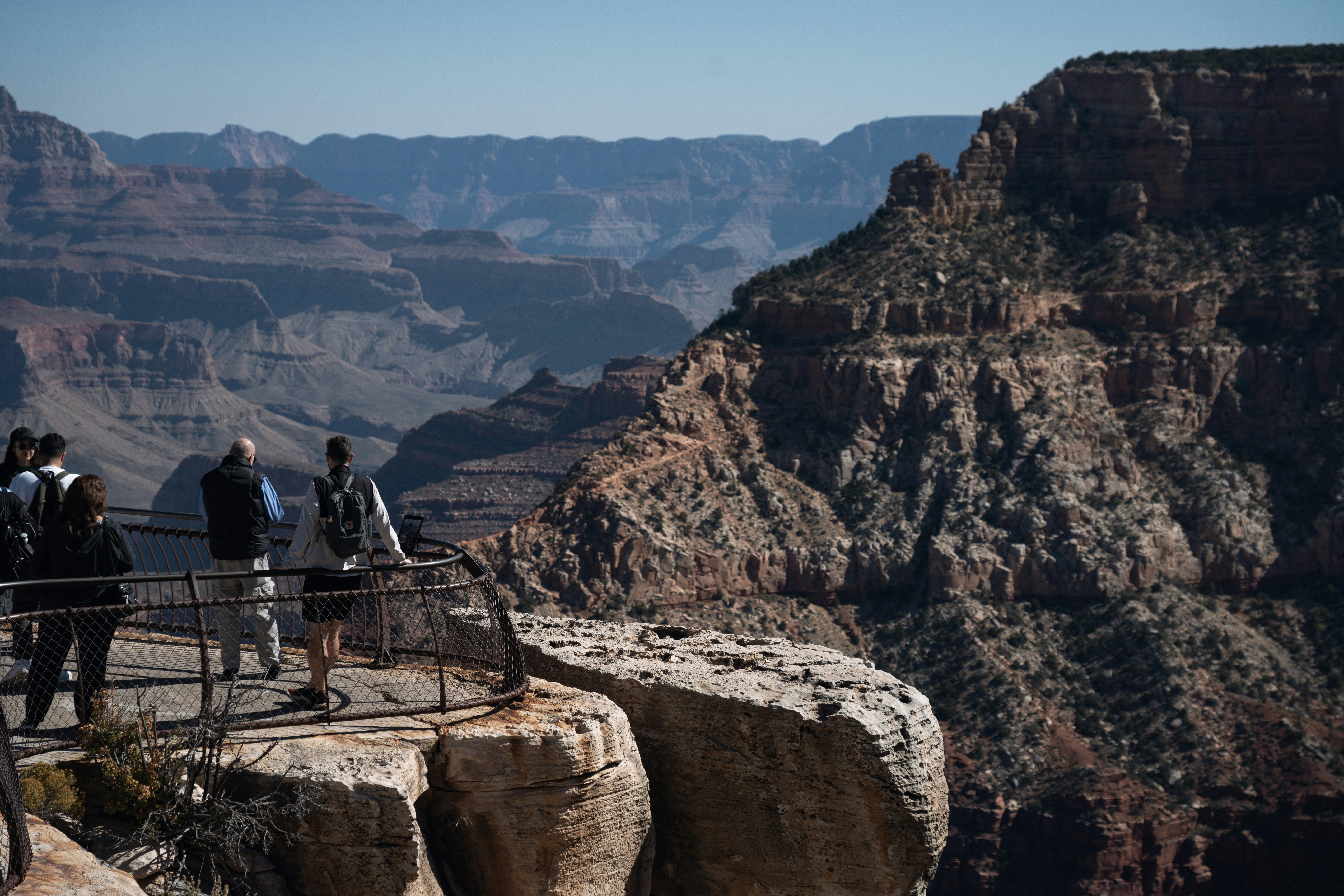 This captivating image captures a group of tourists gazing over the vast expanse of the Grand Canyon from a rocky overlook. The composition highlights the majestic layers of the canyon, ranging from deep reds to subtle browns, under the clear blue sky. The interplay of sunlight and shadow across the rugged landscape enhances the dramatic atmosphere, making the scene visually striking and immersive.