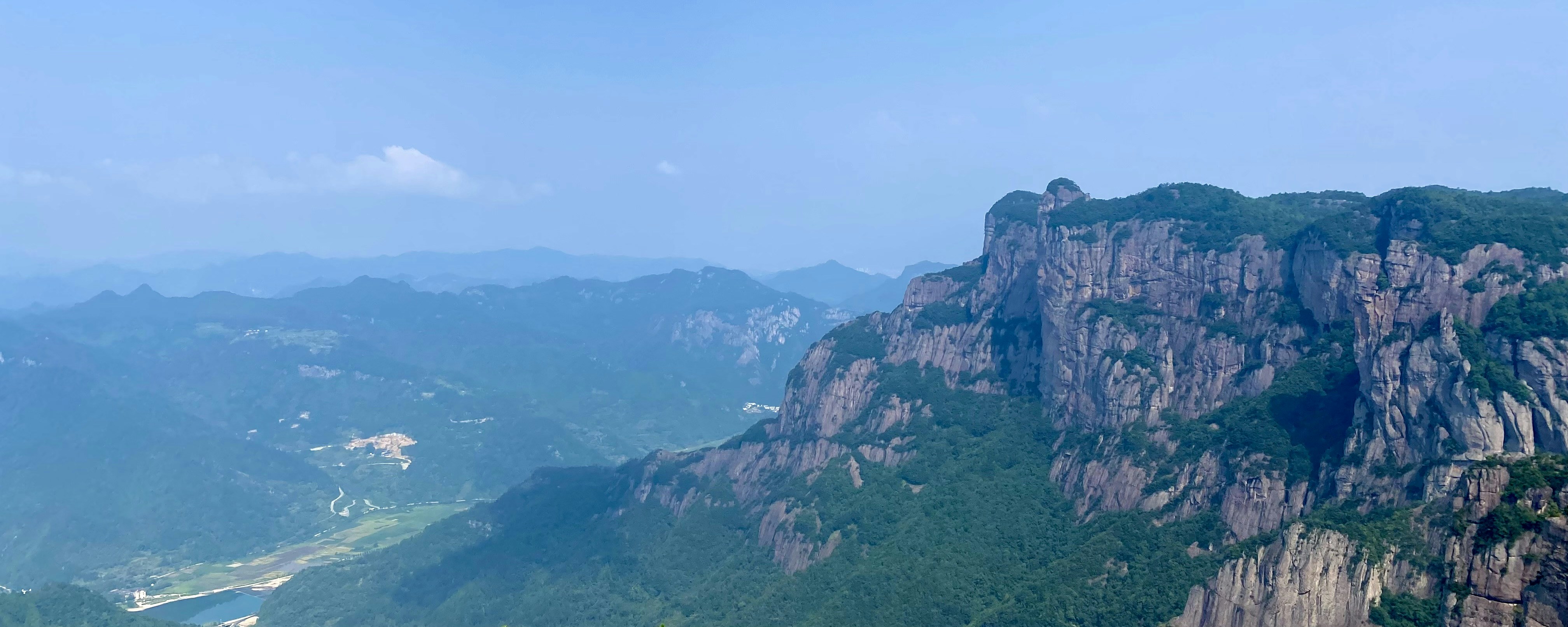 Expansive mountain range viewed from above, with rugged cliffs and lush greenery under a clear blue sky.