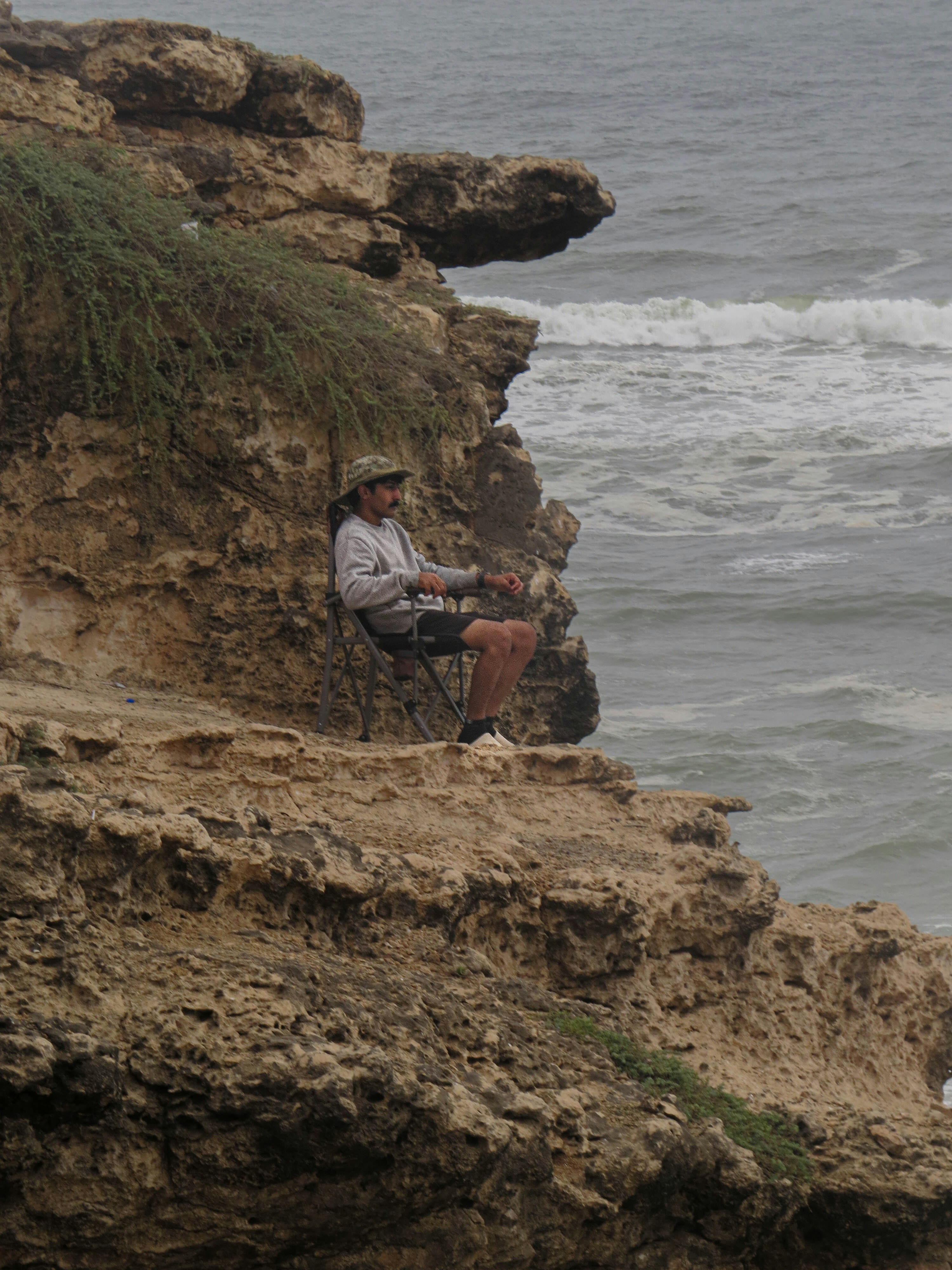 Person seated on rocky cliff overlooking the ocean waves under an overcast sky.
