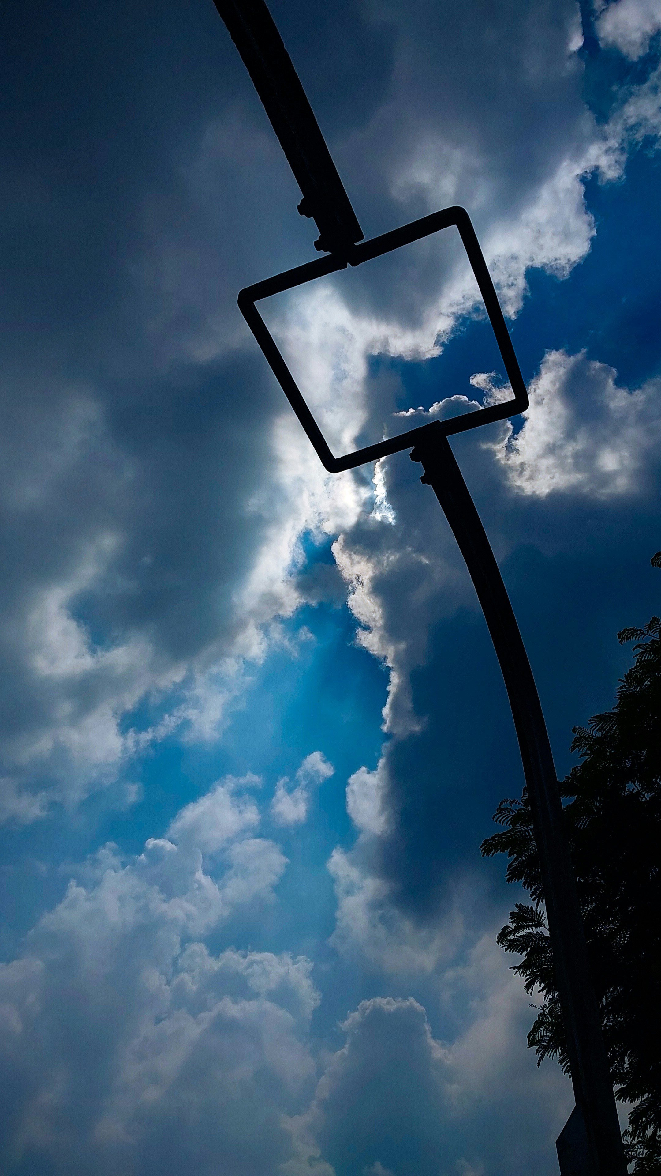 Silhouetted square metal frame mounted on a curved pole rises against a dramatic, cloud-filled sky. The composition emphasizes geometric contrast between industrial steel and the expansive blue atmosphere.