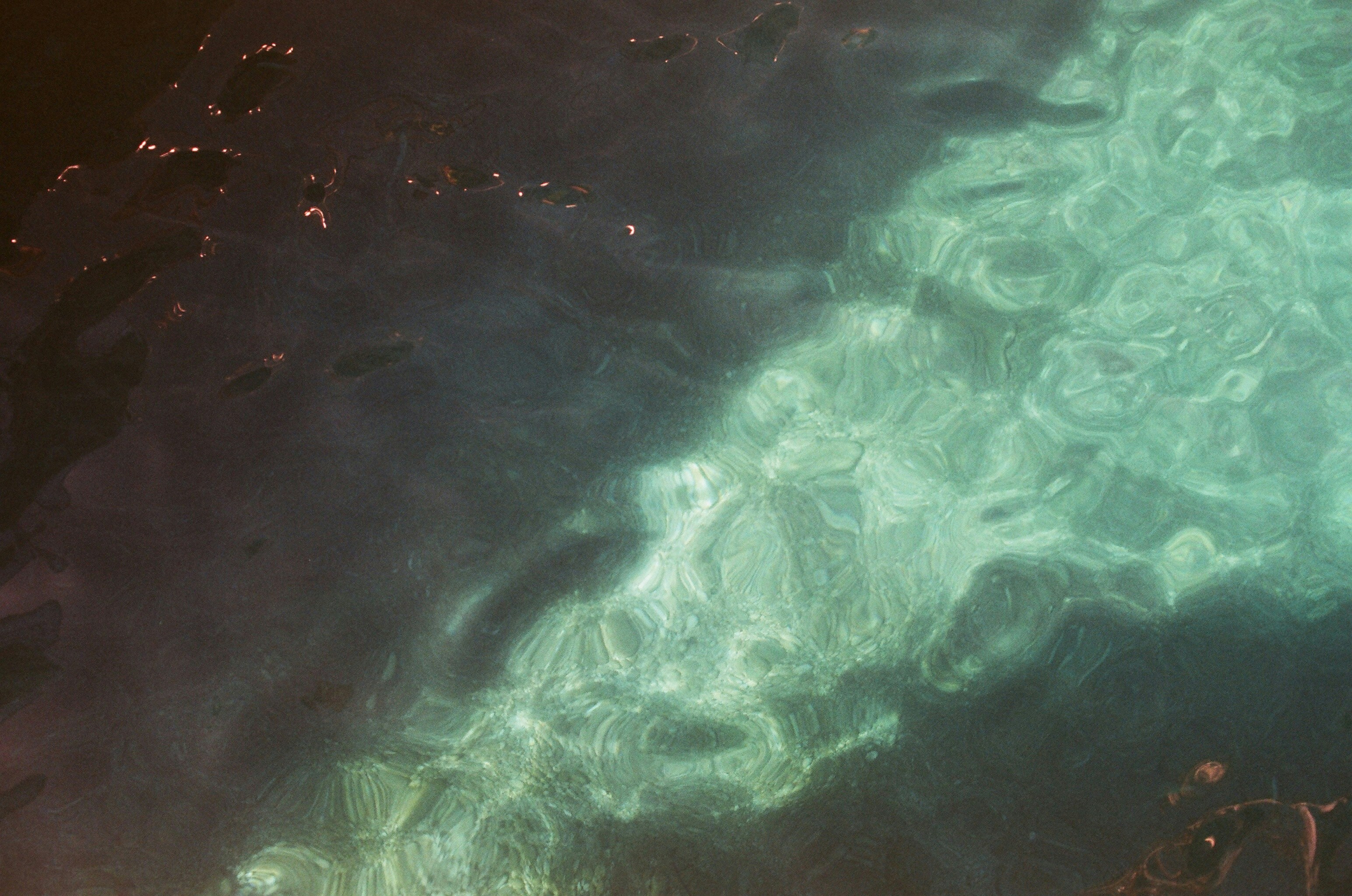 Sunlit turquoise water reveals intricate ripples along a shadowed shoreline.