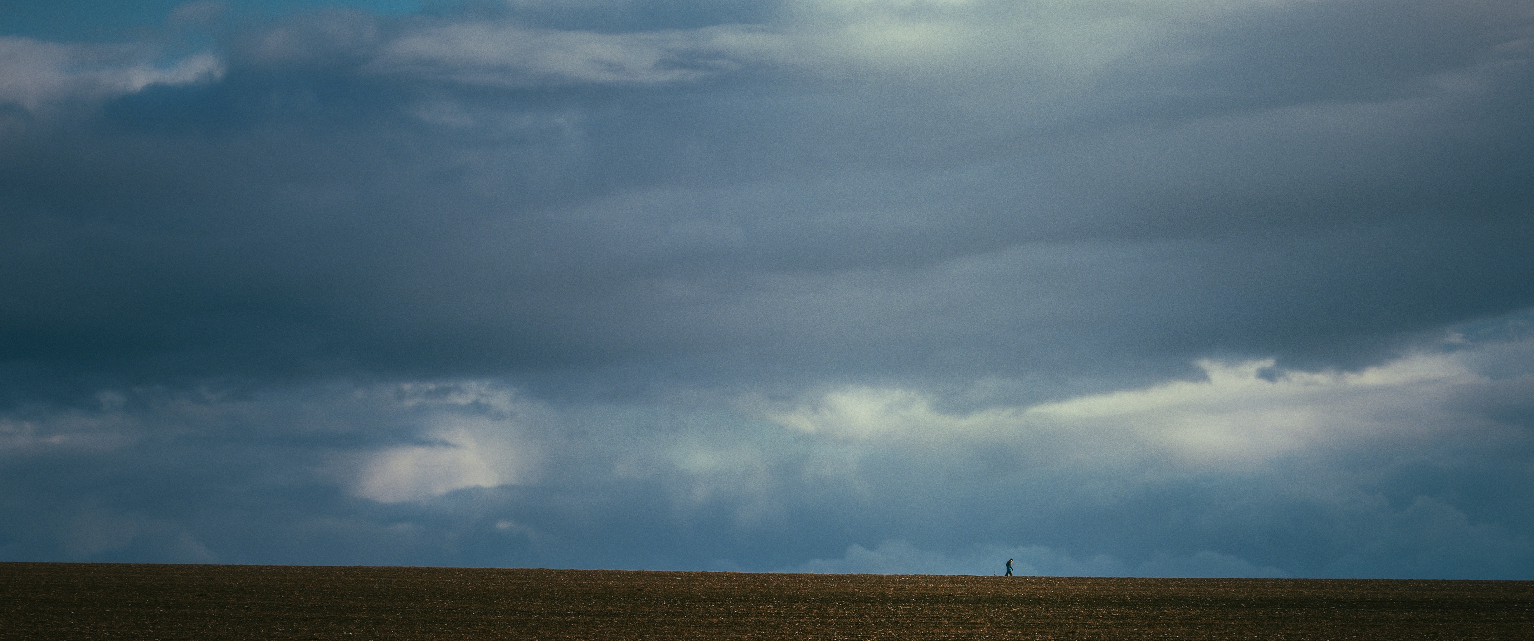 Lone figure walking along a vast landscape beneath dramatic cloud cover.