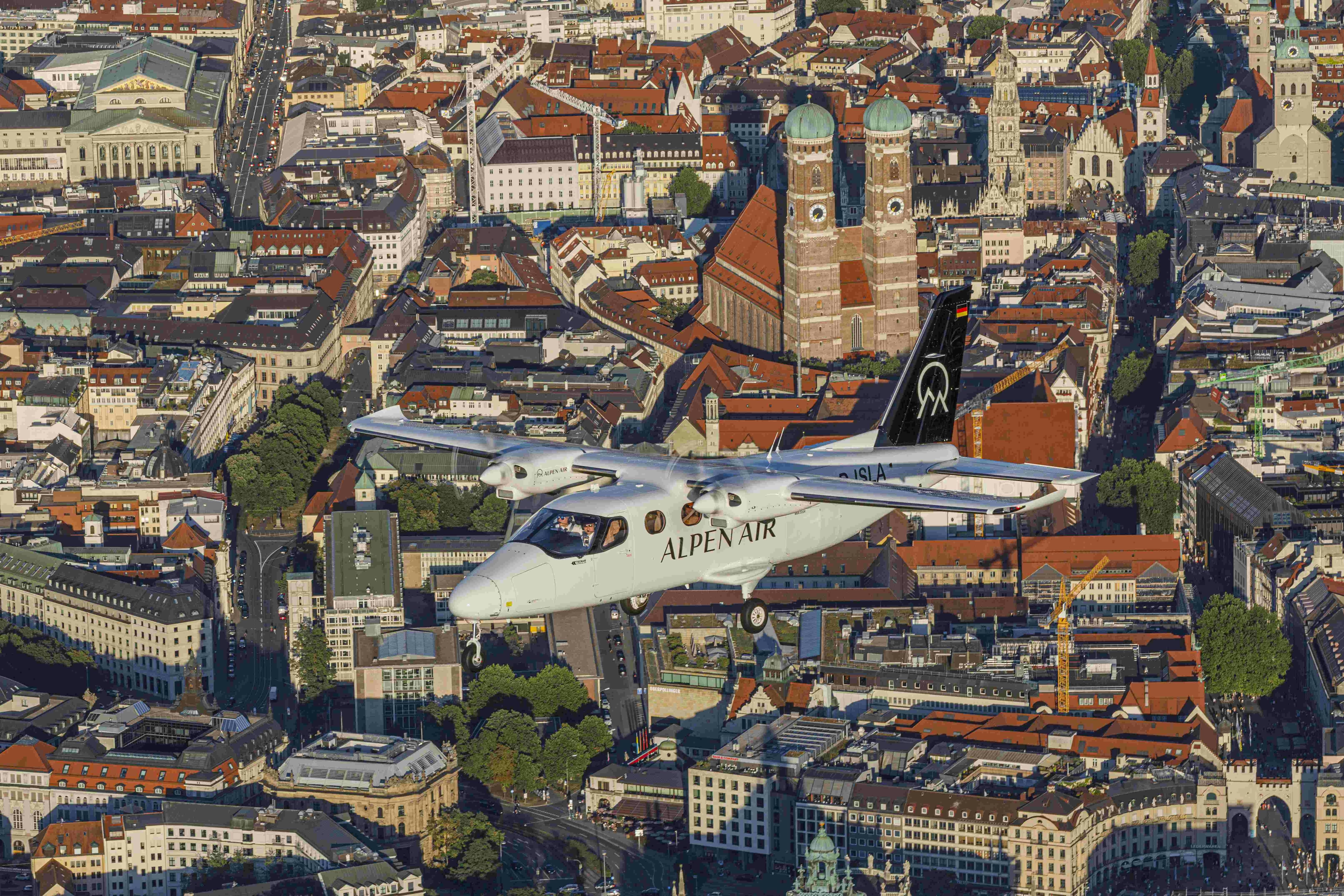 The Alpen Air´s Tecnam P2012 Traveller aircraft above the Bavarian city of Munich with houses, big buildings and the famous church Frauenkirche. Two pilots can be seen smiling in the cockpit.