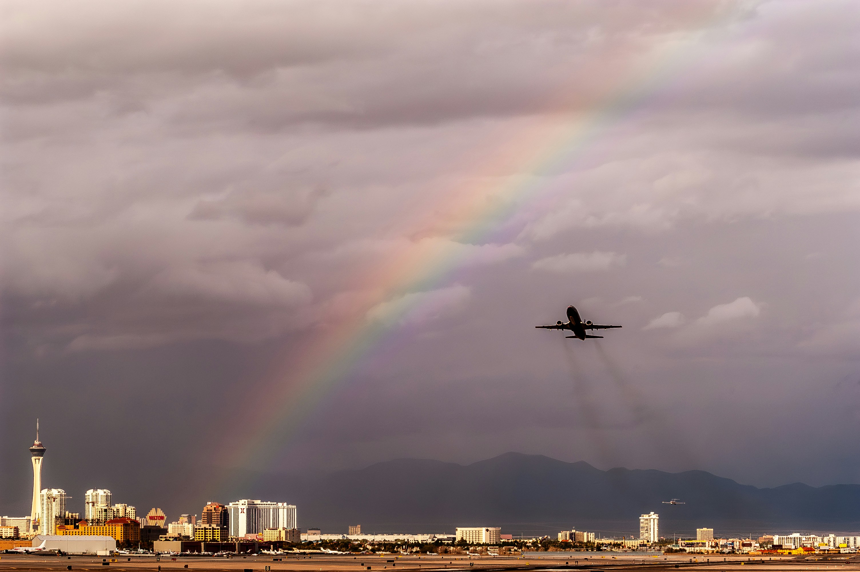 Airplane ascending with a vibrant rainbow arching over the Las Vegas skyline.
