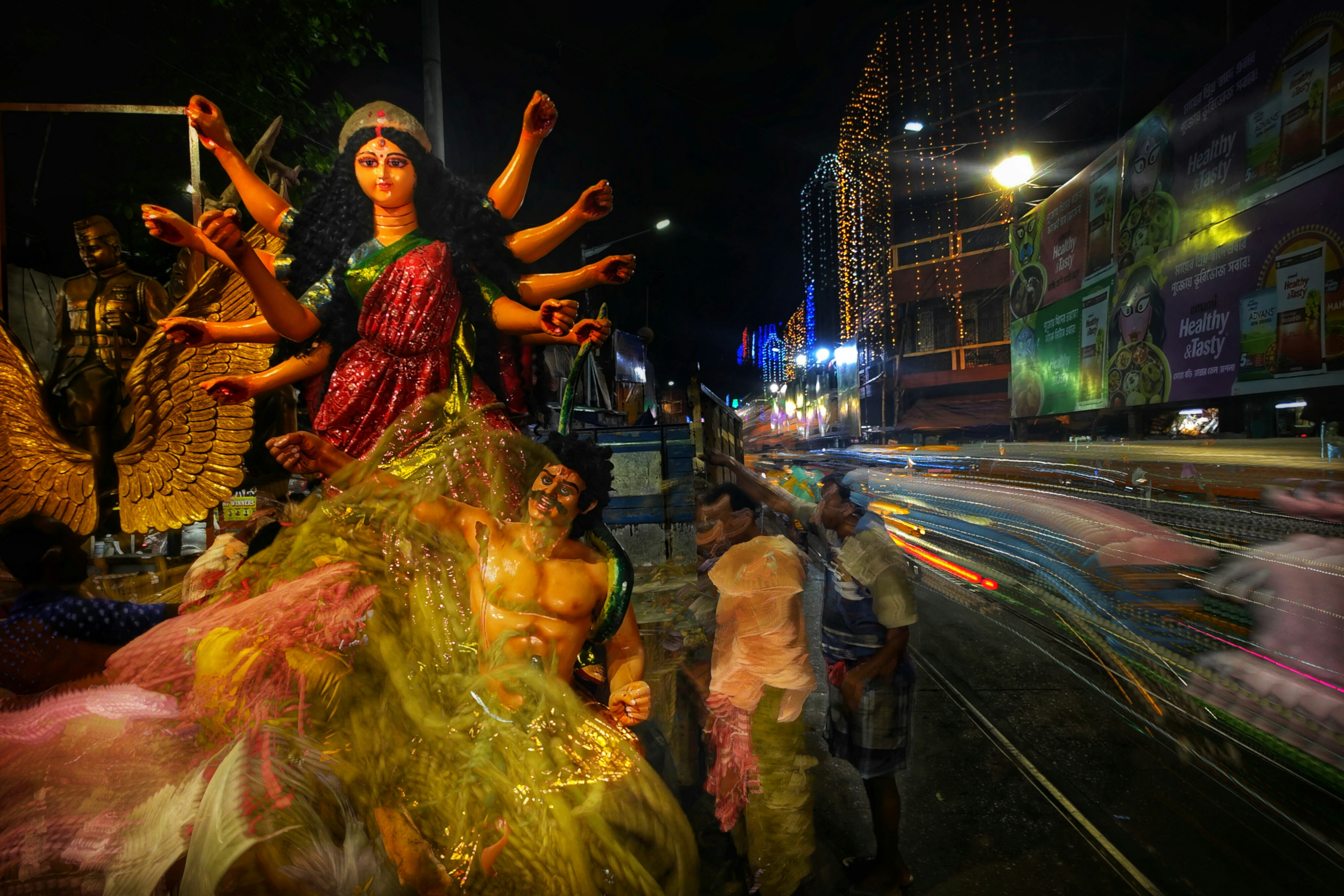 Night-time photograph of a multi-armed Durga idol in a street procession, surrounded by vibrant fabrics and streaks of light from passing traffic.