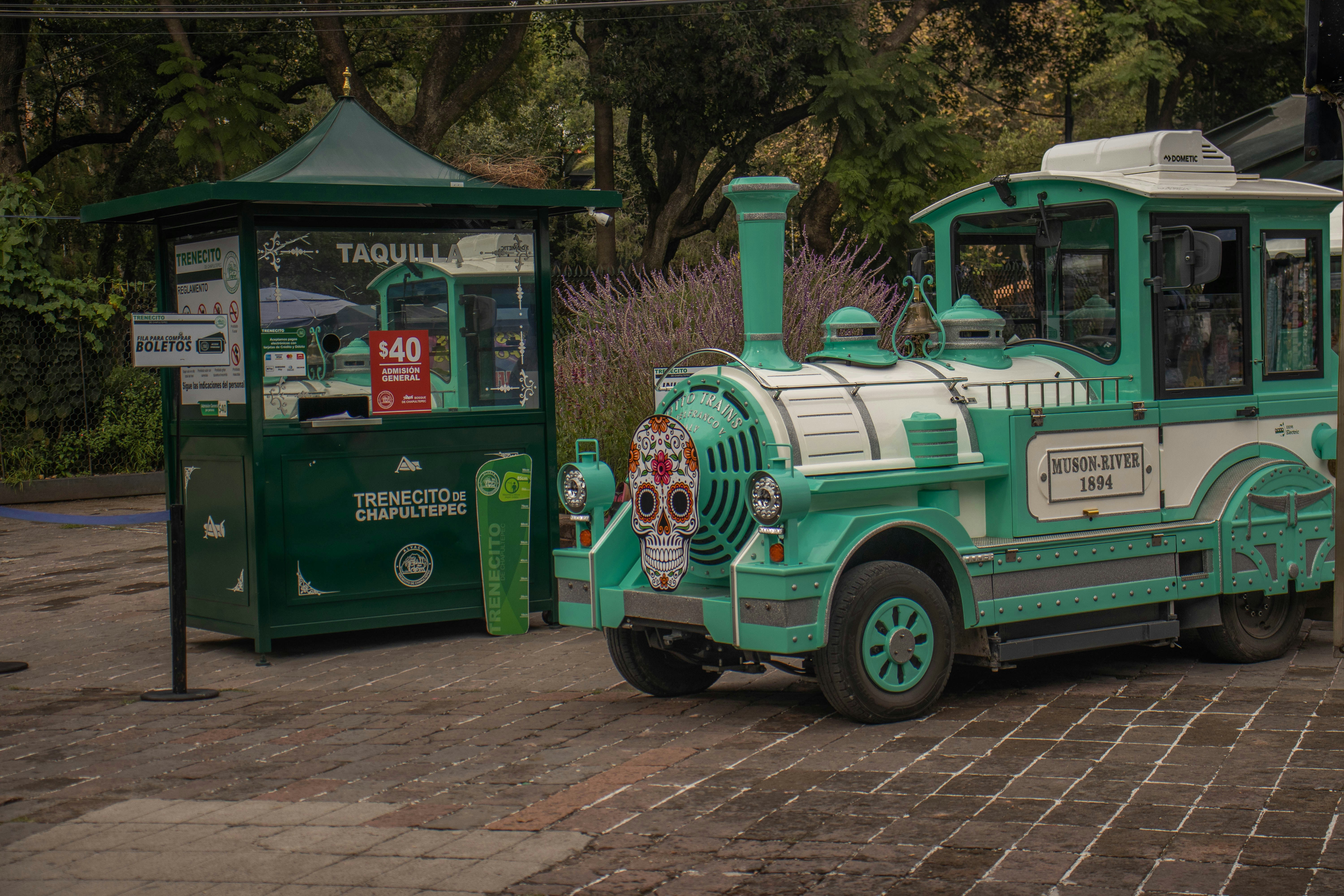 Green and white vintage-style train parked next to a ticket booth in a leafy park setting.