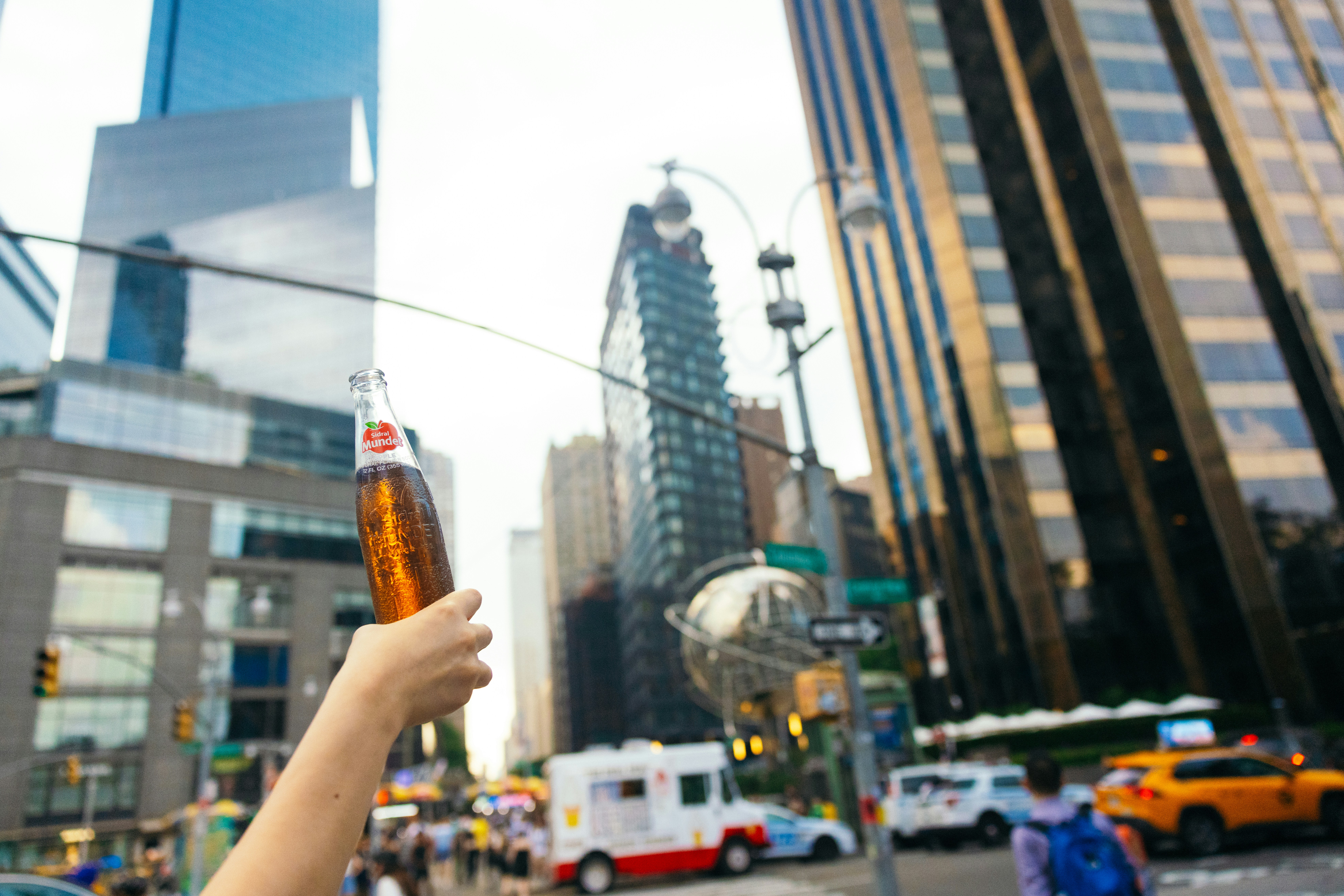 A hand raises a soda bottle in the foreground while a bustling city street with tall skyscrapers unfolds in the background. The shot emphasizes urban scale and motion.