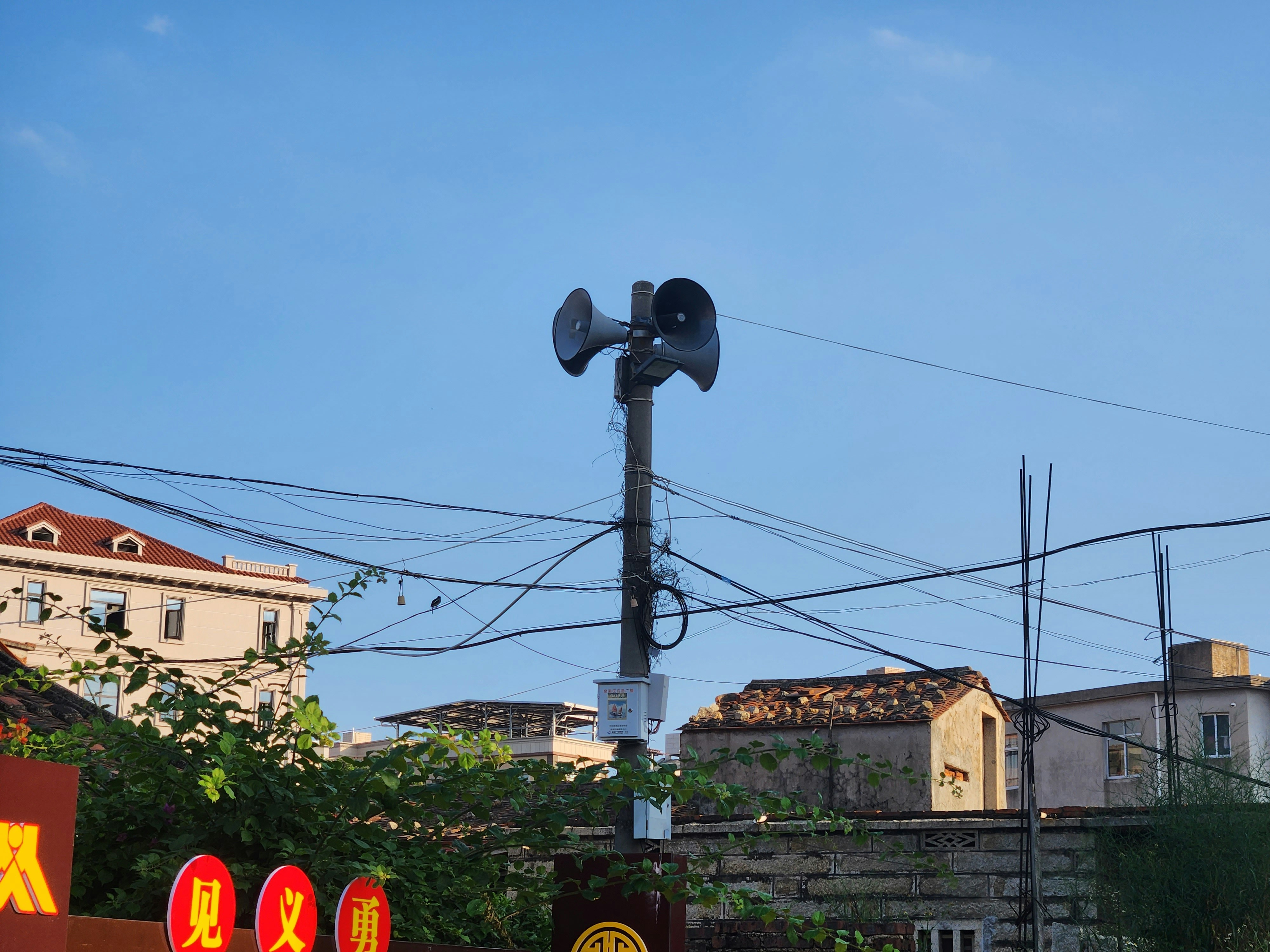 A street pole bears a dual megaphone on top, rising above a network of wires. Colorful storefront signs and weathered buildings anchor the lower frame under a clear blue sky.