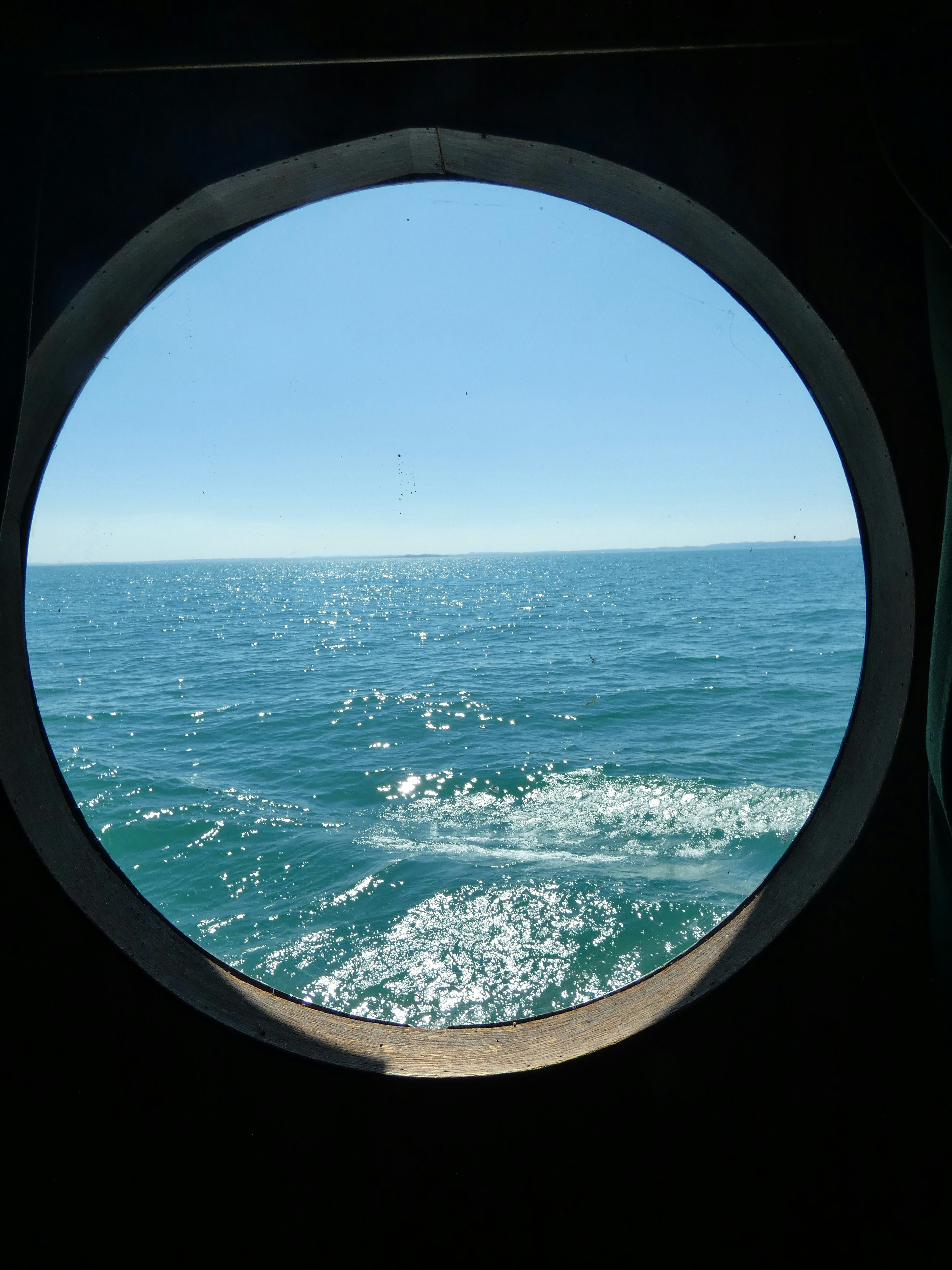 Photograph of a sunlit blue ocean framed by a circular porthole, with glittering waves along the horizon. The composition emphasizes the contrast between the dark interior and the bright seascape.