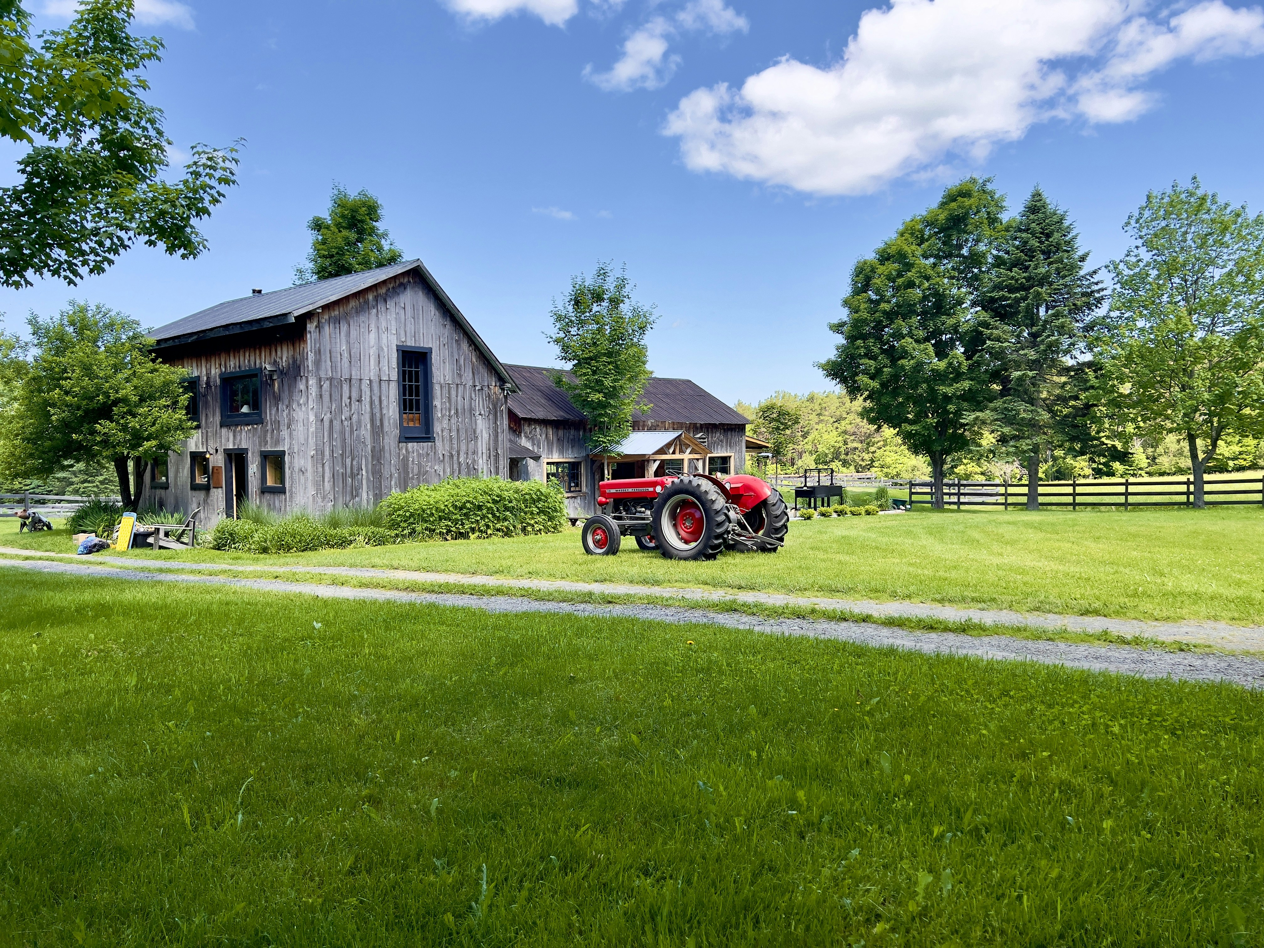 A vintage red tractor rests in a vibrant green field beside a rustic wooden barn, framed by lush trees under a clear blue sky.