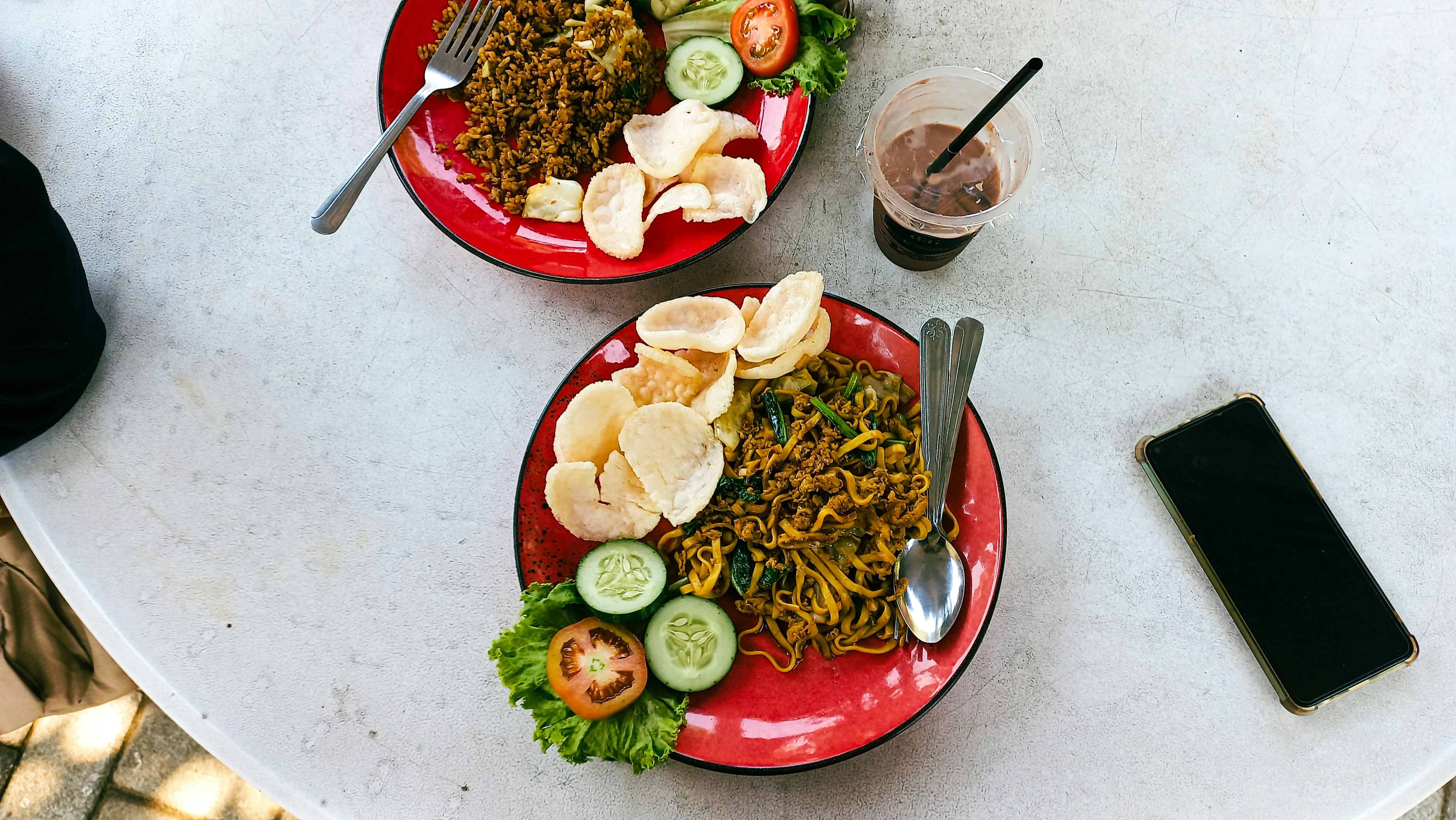 Two red plates of noodles with sliced cucumbers, tomatoes, and crispy chips on a white table beside a smartphone.